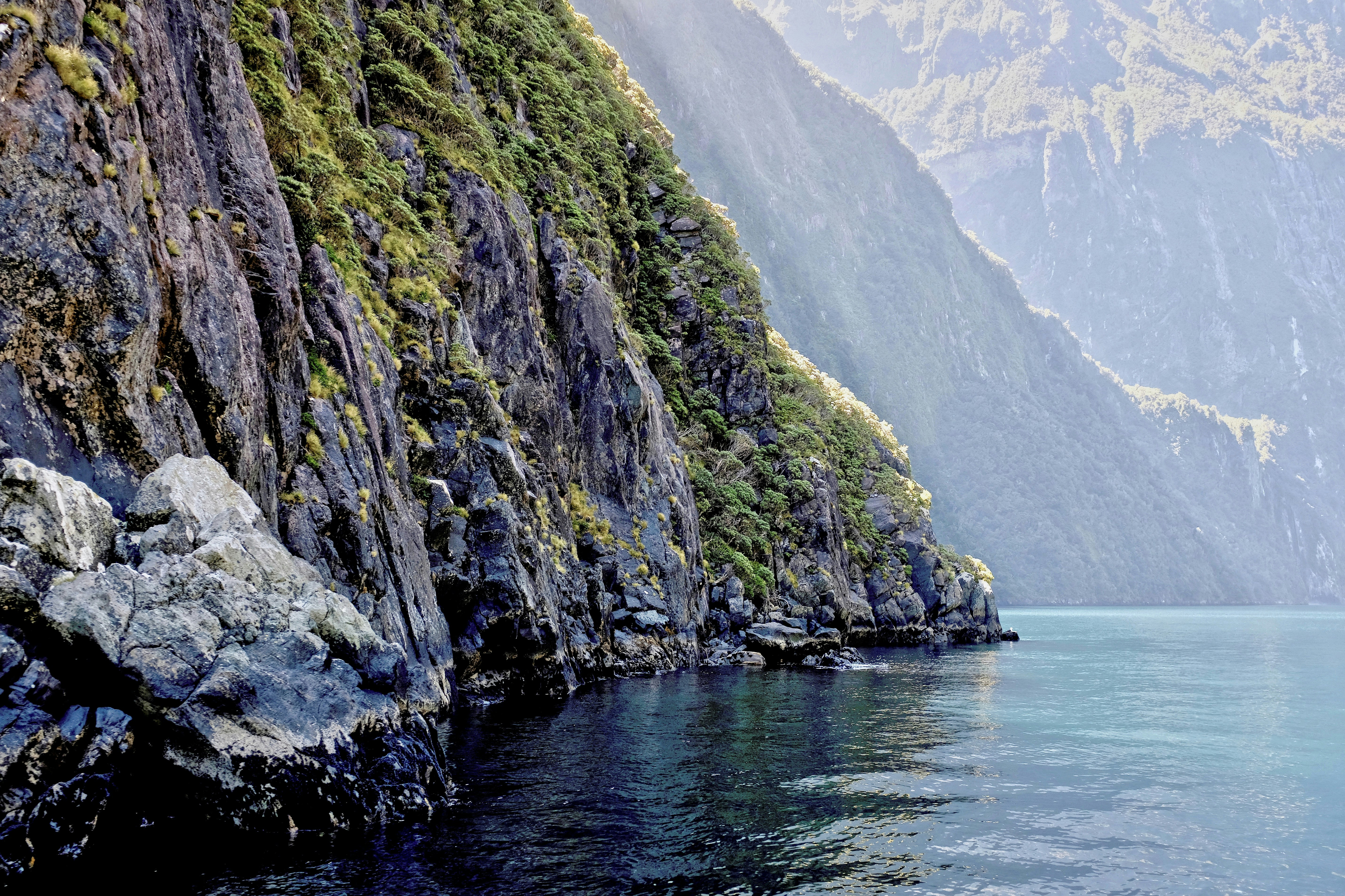 Sunlight gently illuminates steep cliffs covered in lush greenery, casting reflections over tranquil blue waters.