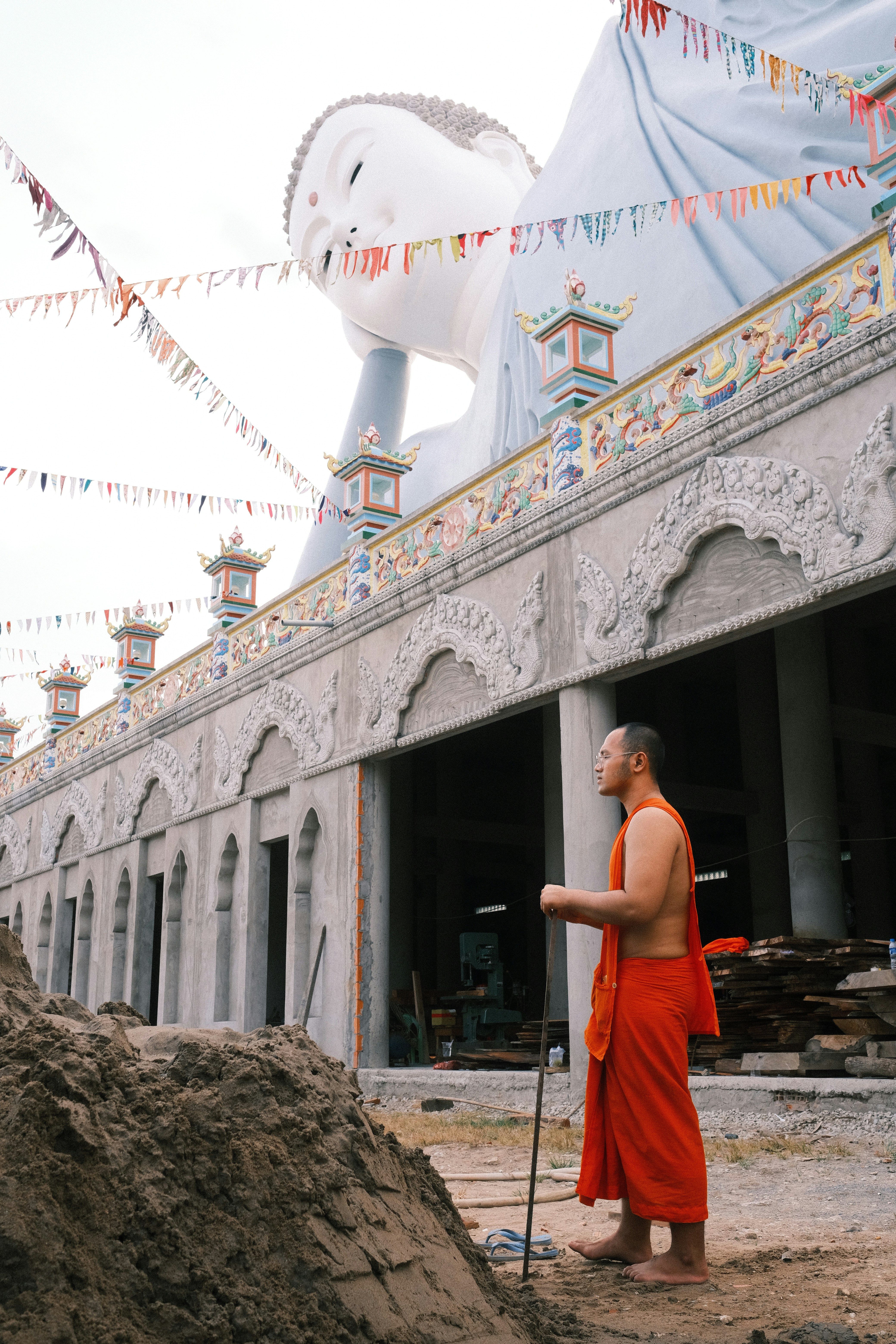 A barefoot monk in an orange robe stands with a staff before an ornate temple facade beneath a colossal white Buddha head and fluttering pennant banners.