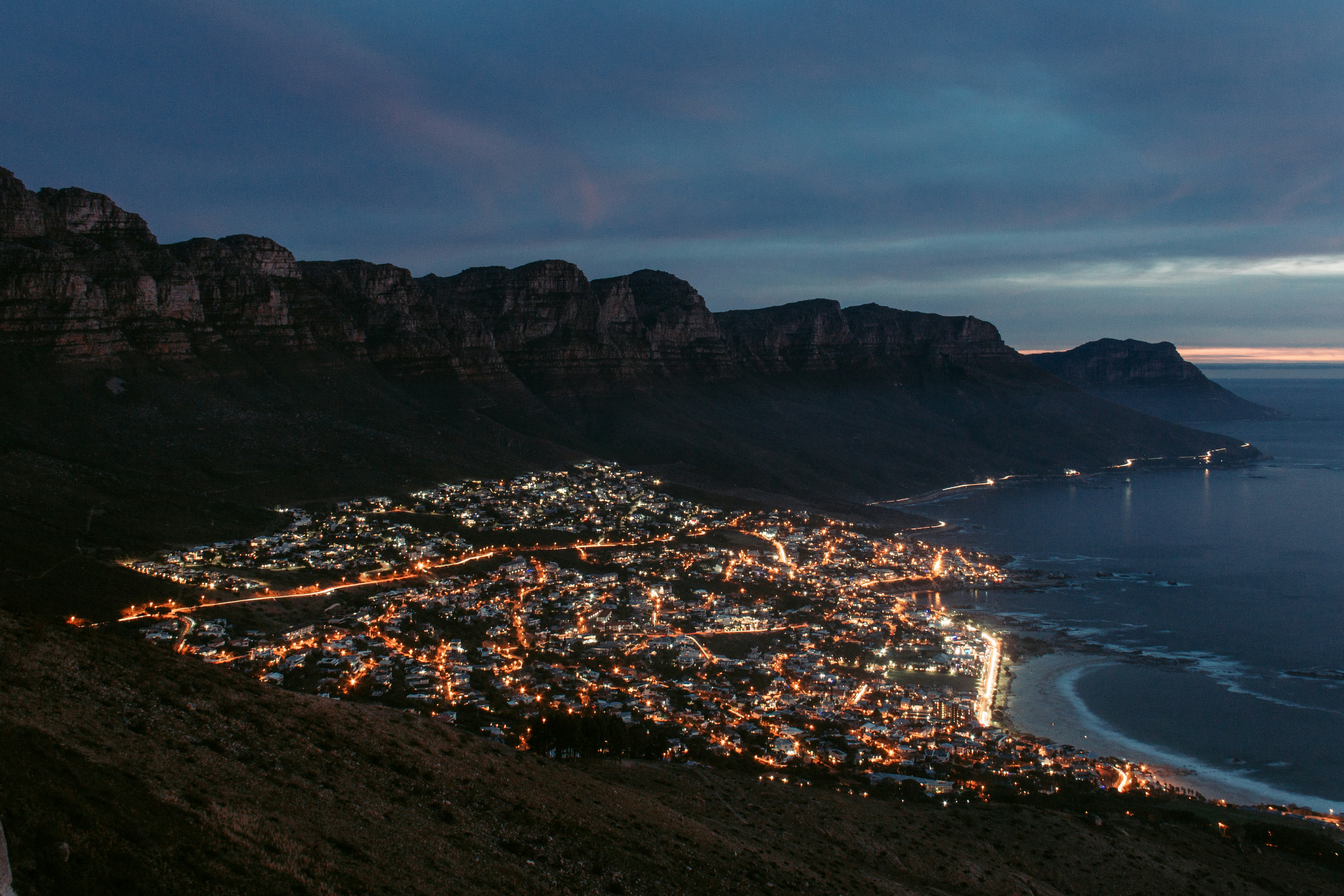 Illuminated coastal town nestled against rugged mountains at twilight, showcasing a blend of natural beauty and urban life.
