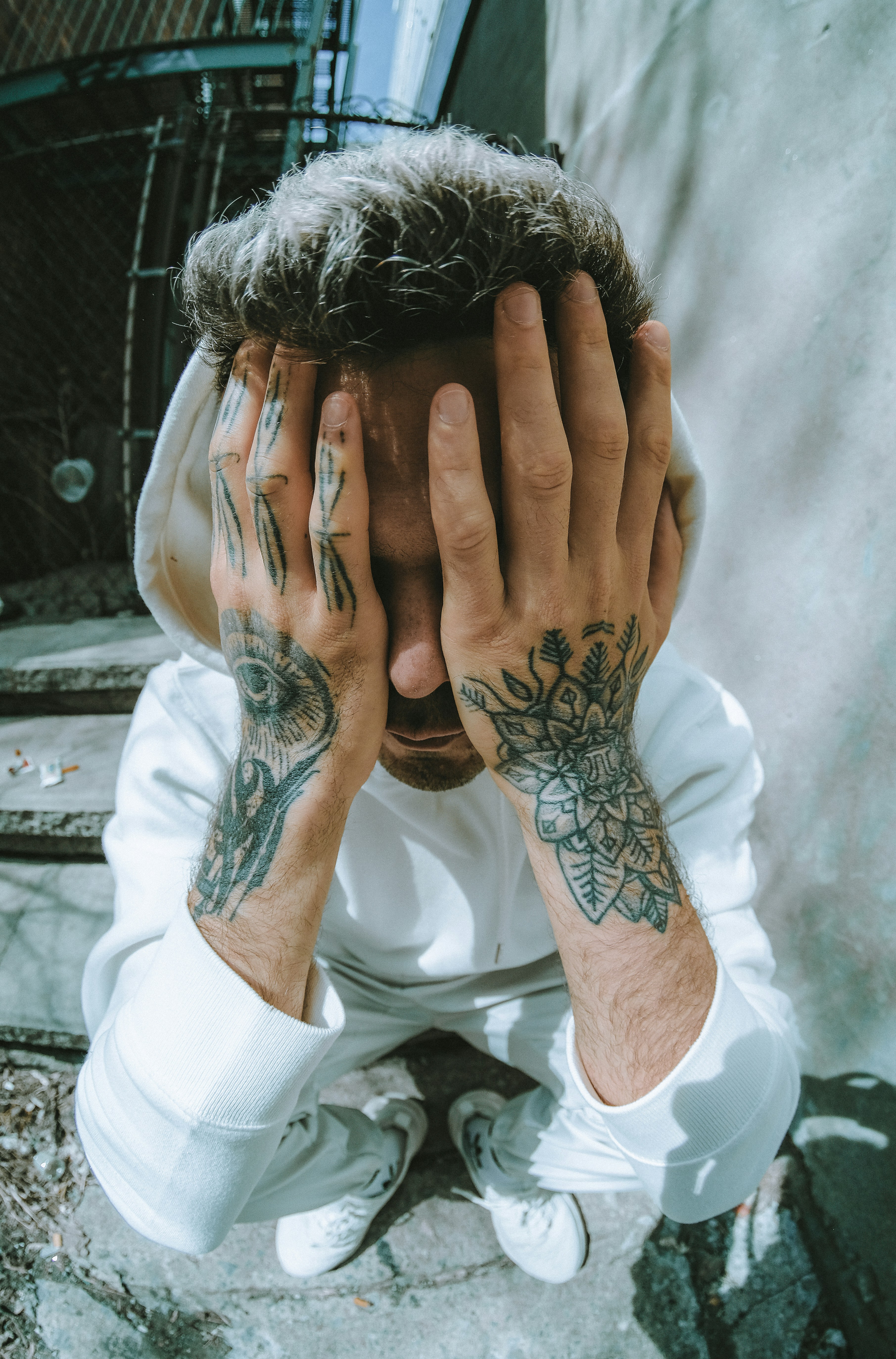 A person in a white hoodie sits on stairs, covering their face with tattooed hands, conveying a sense of introspection and vulnerability.
