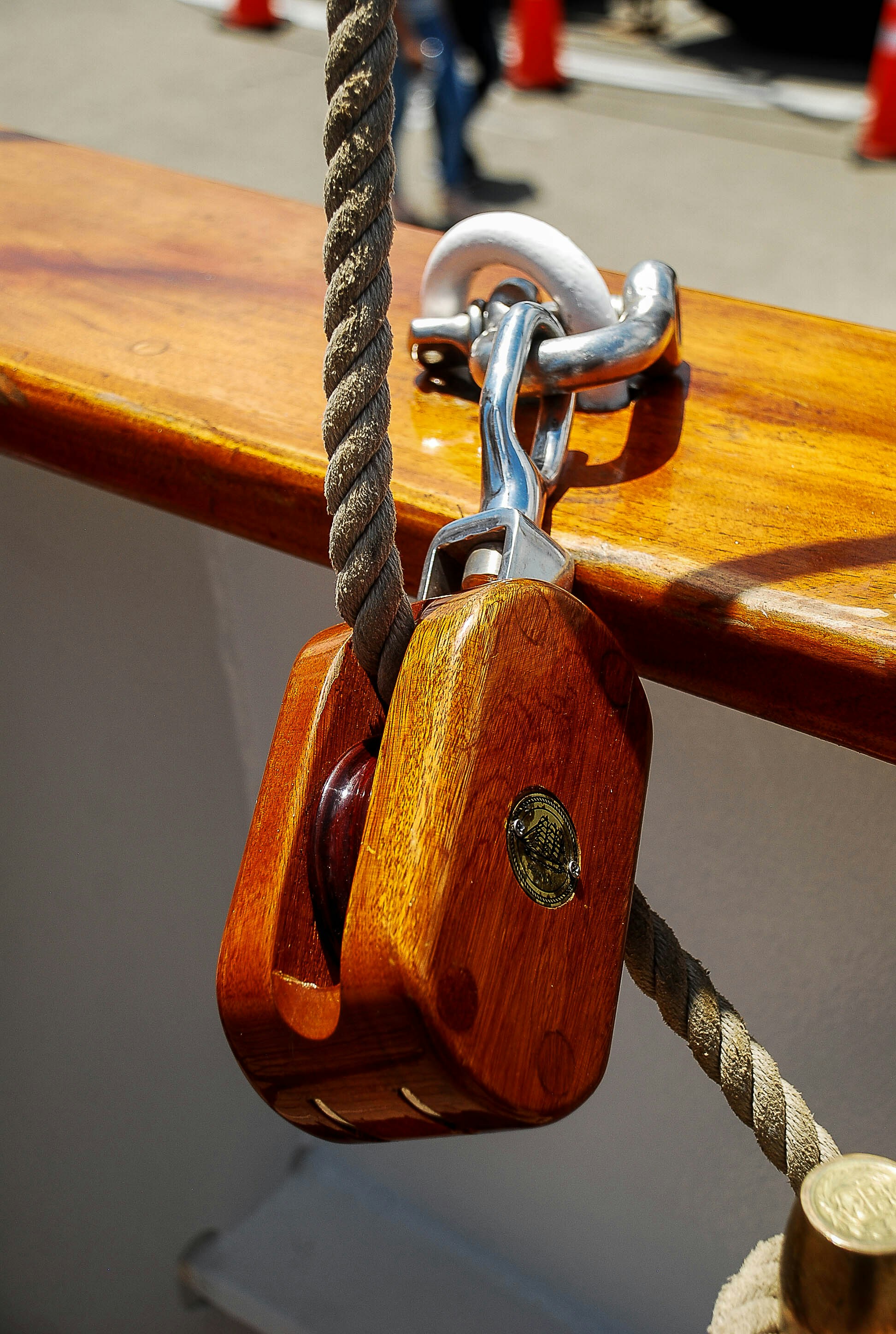 Close-up of a polished wooden pulley attached to a rope, showcasing intricate details and craftsmanship. The background features blurred elements of a maritime setting.