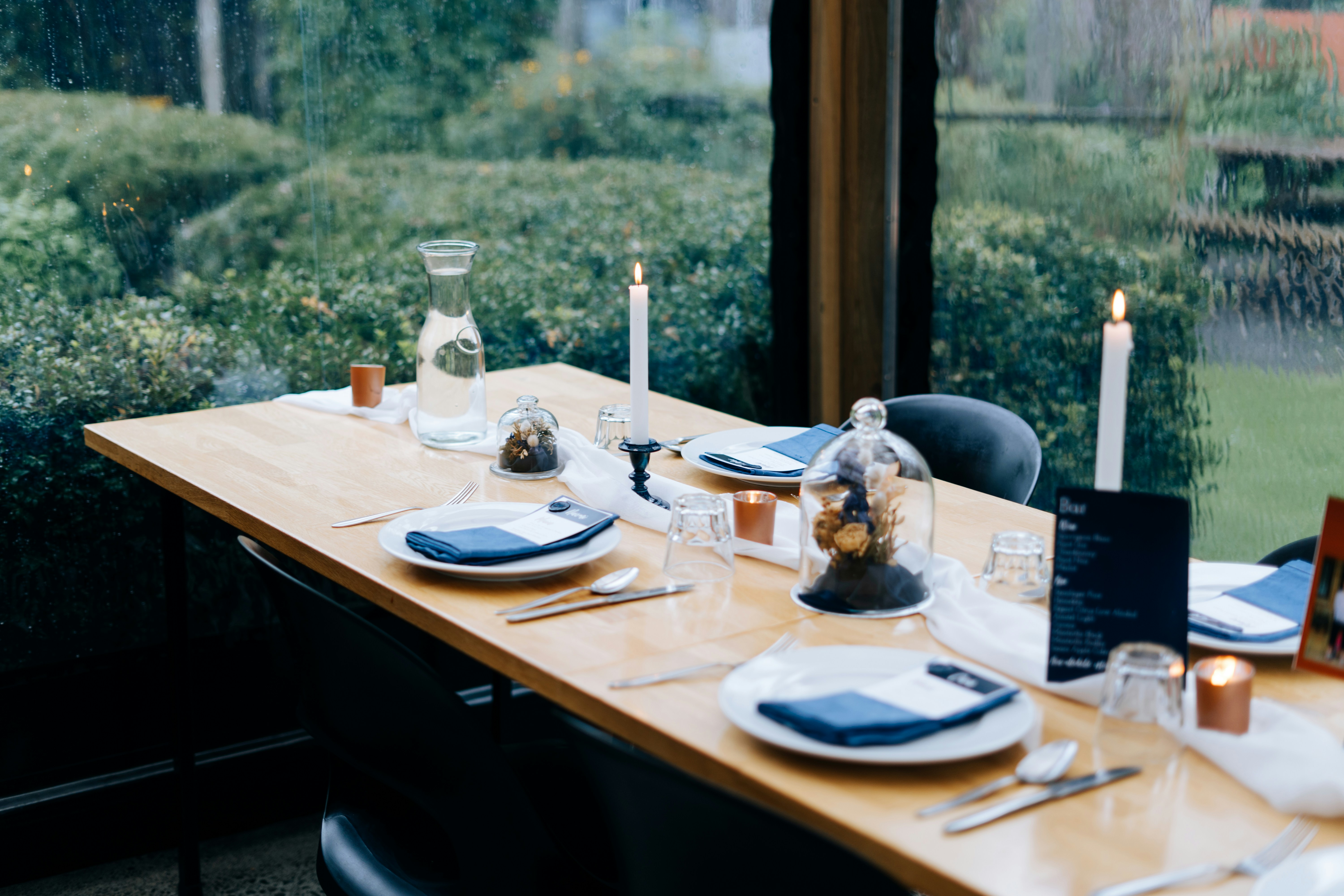 Elegantly set dining table with candles and decorative elements, framed by a backdrop of lush greenery and rain-streaked glass.