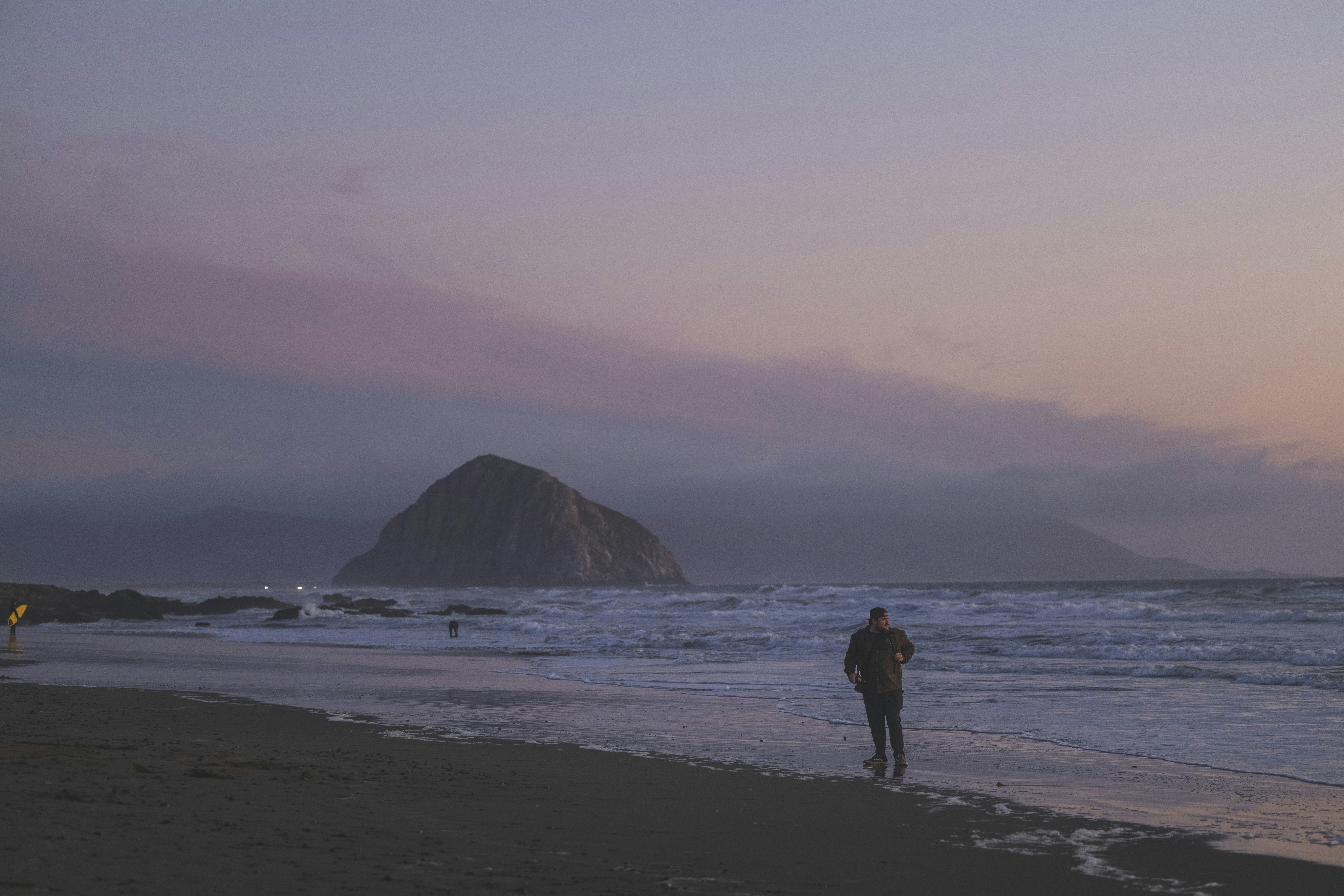 A lone figure walks along the shoreline, with a prominent rock formation in the background under a twilight sky.