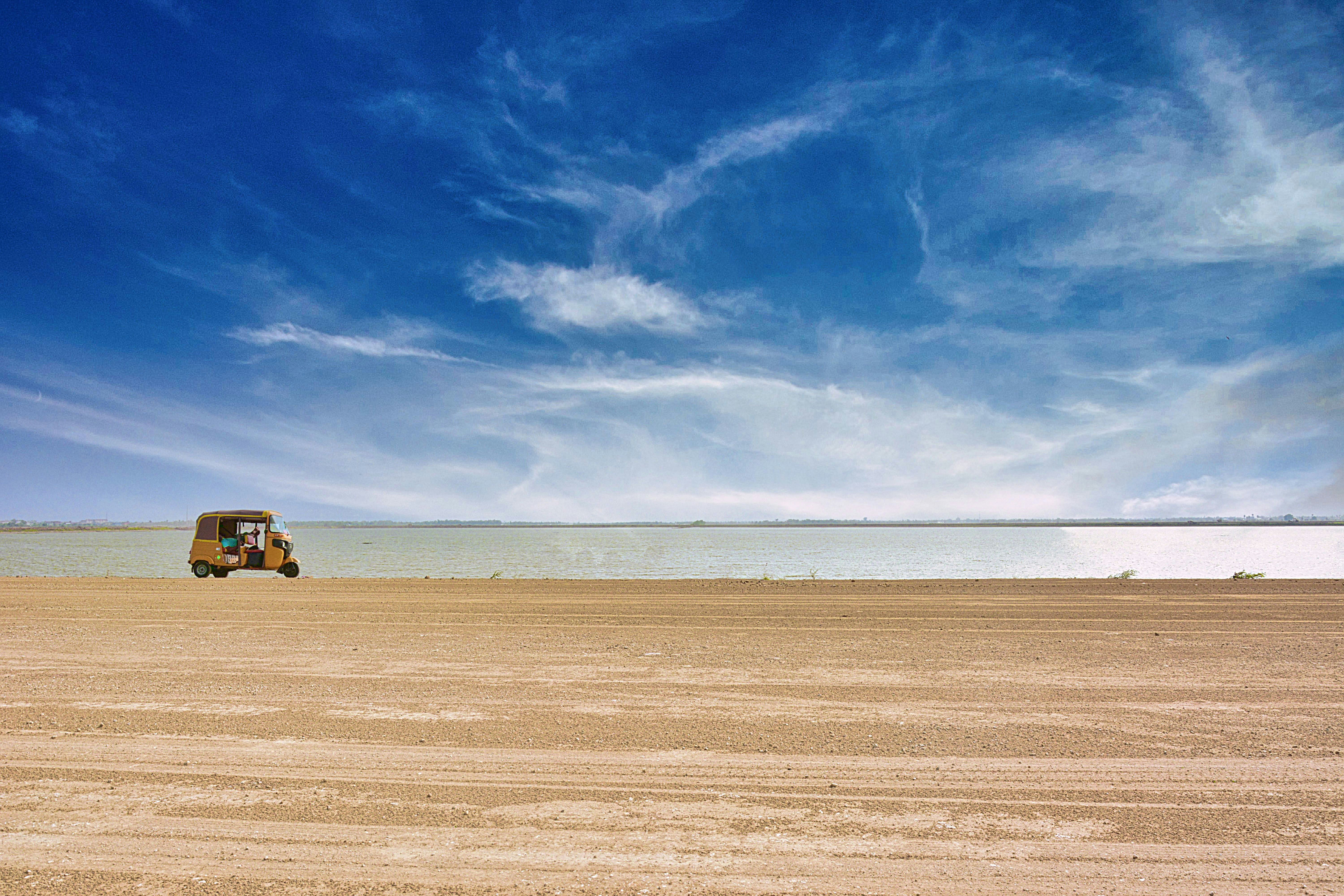 An auto rickshaw navigates a vast, sandy landscape under an expansive blue sky, reflecting a serene journey by the water's edge.
