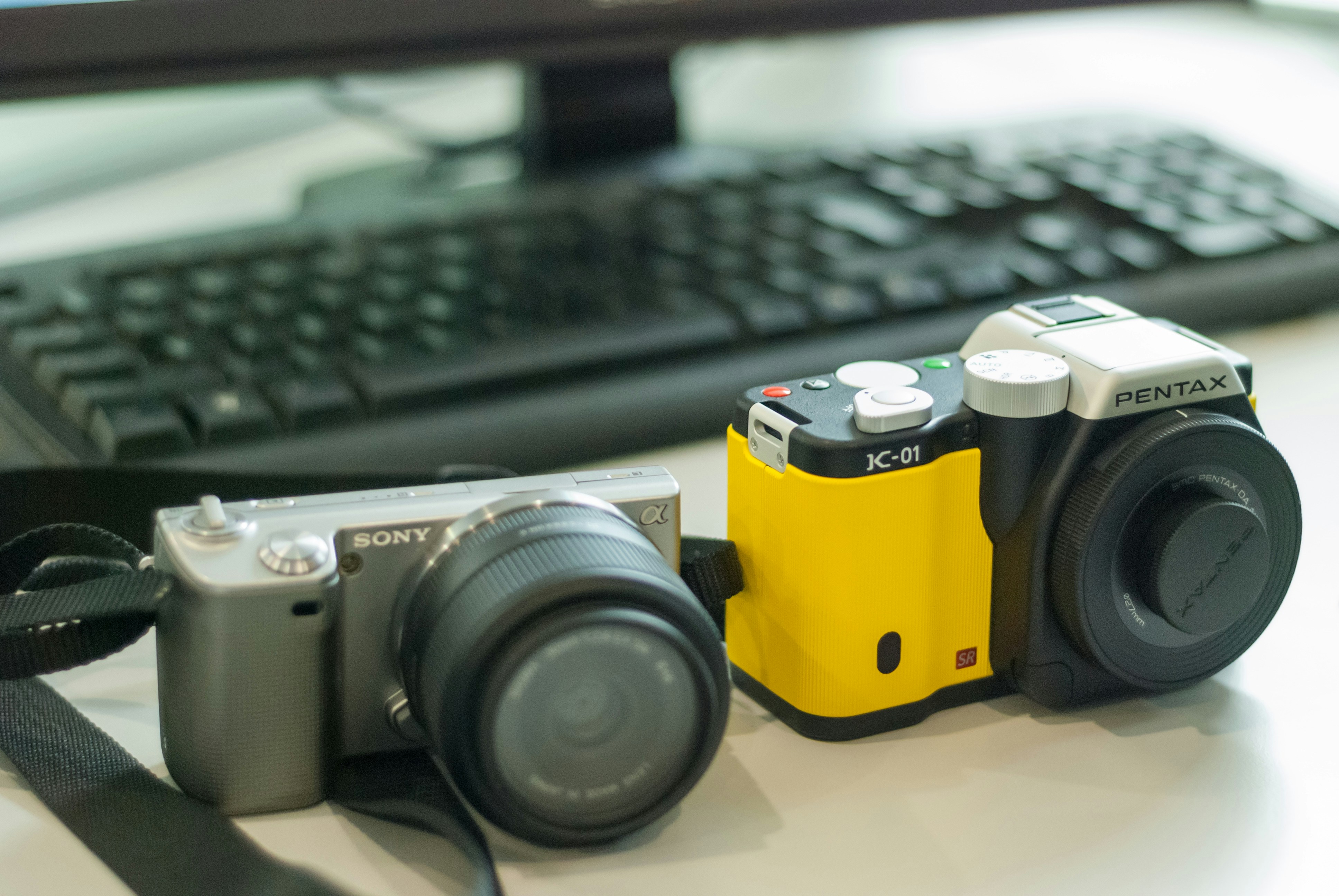Sunlit Yellow Pentax JC-01 Flanked by Silver Sony on a Desk