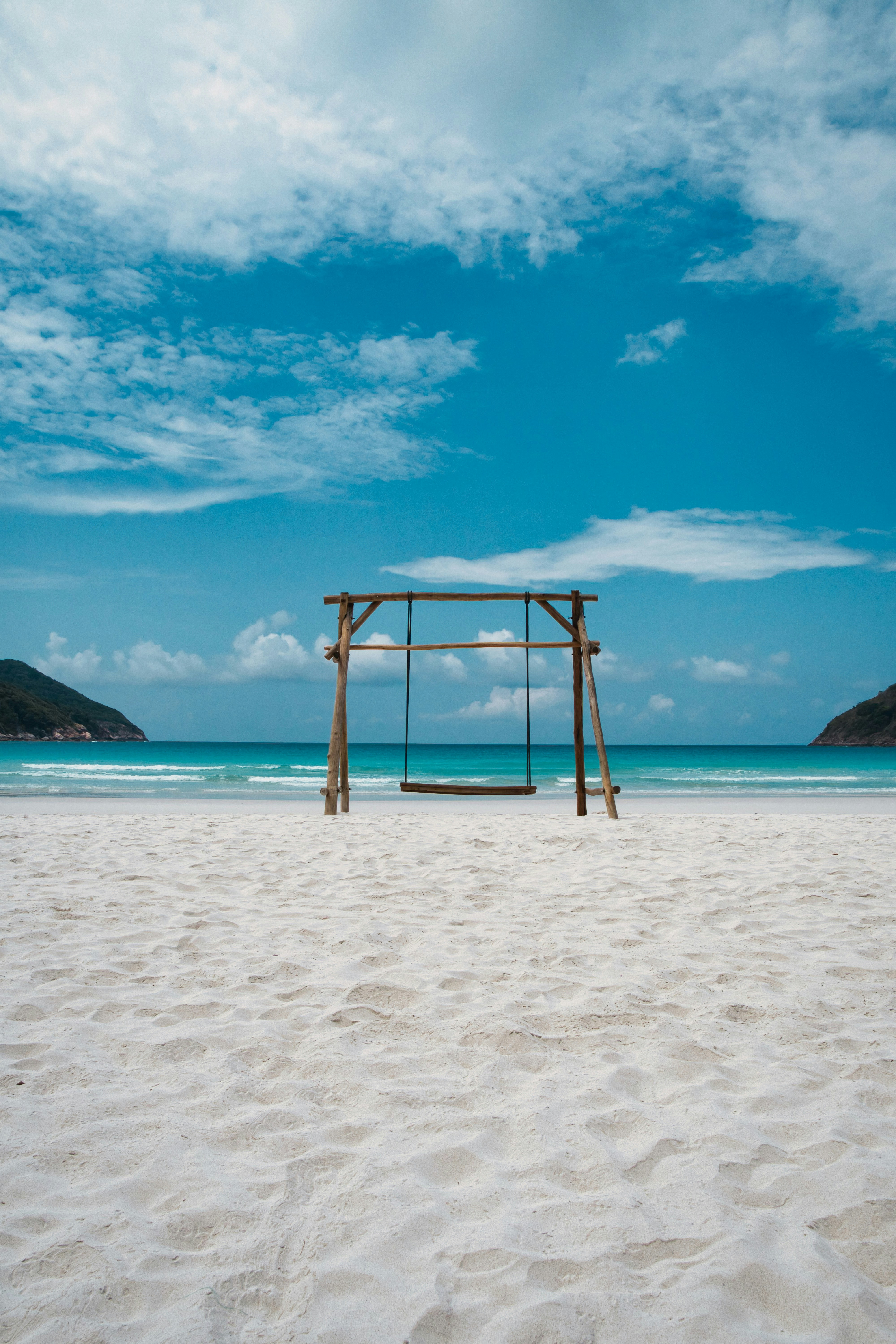 Wooden swing hanging from a rustic frame on a pristine beach, framed by calm turquoise waters and a clear blue sky.