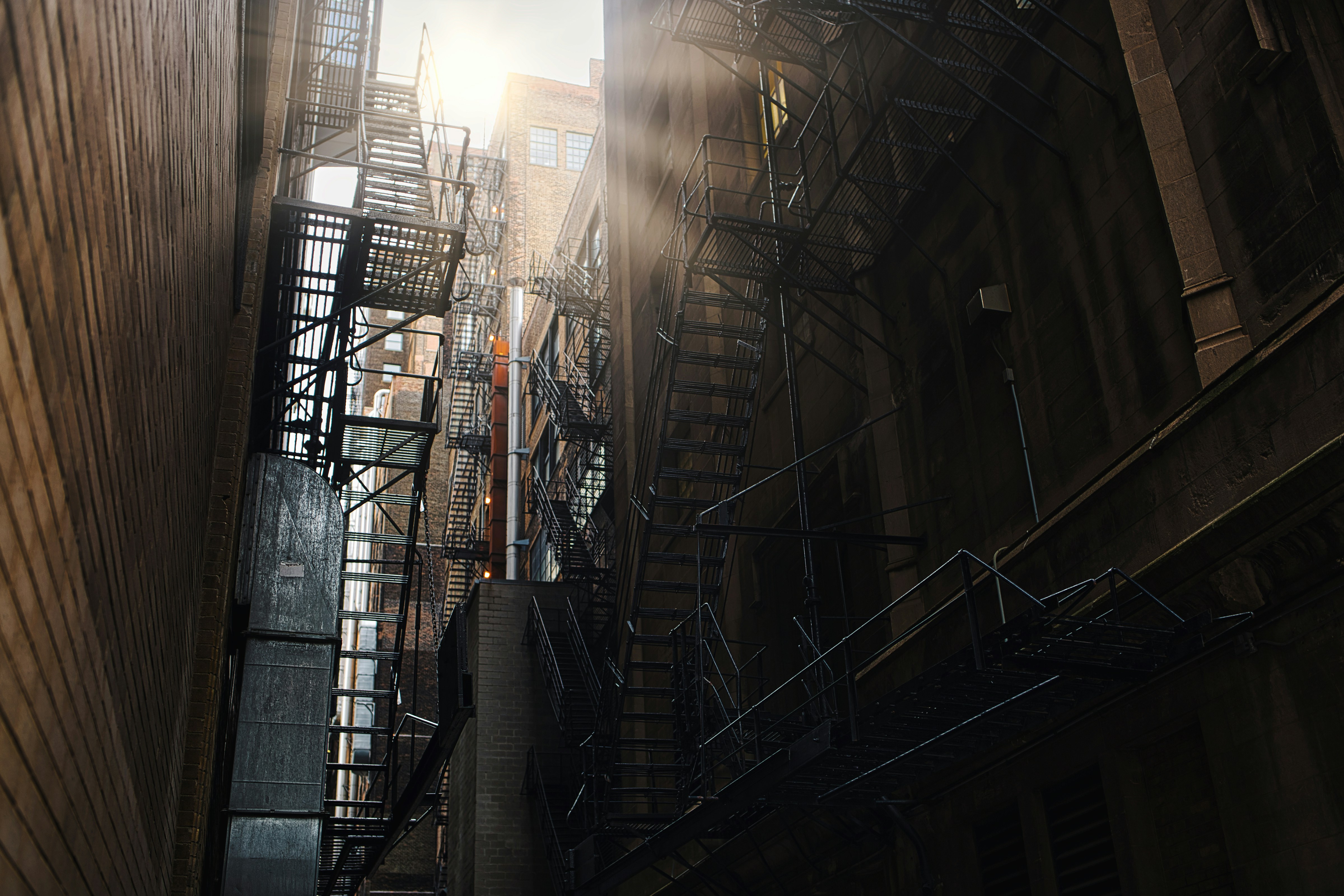 Narrow alleyway with fire escapes and sunlight filtering through buildings, highlighting the contrast between urban structures and natural light.