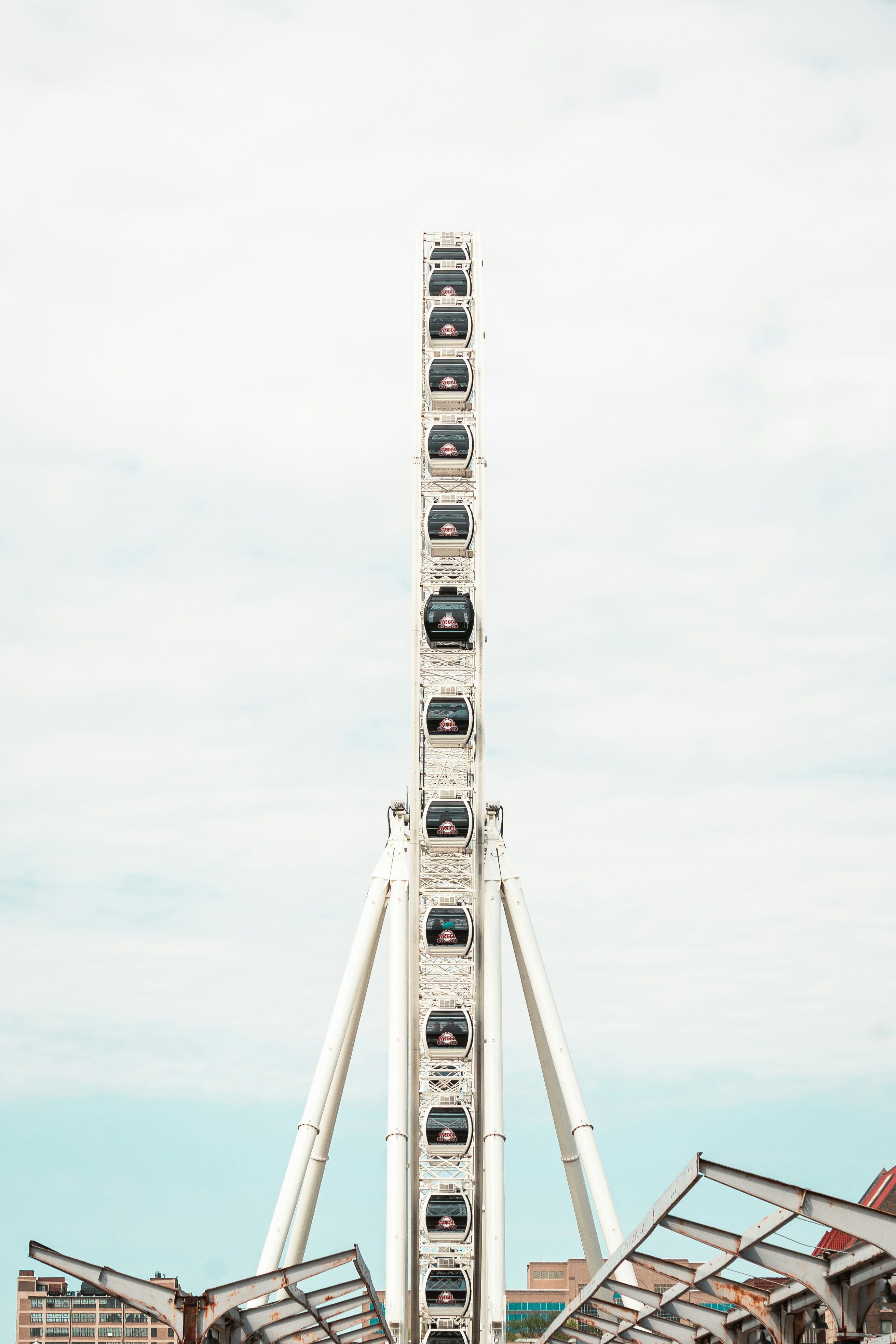 A towering Ferris wheel stands against a pale sky, showcasing its passenger cabins suspended in mid-air. The structure's sleek design contrasts with the urban backdrop.