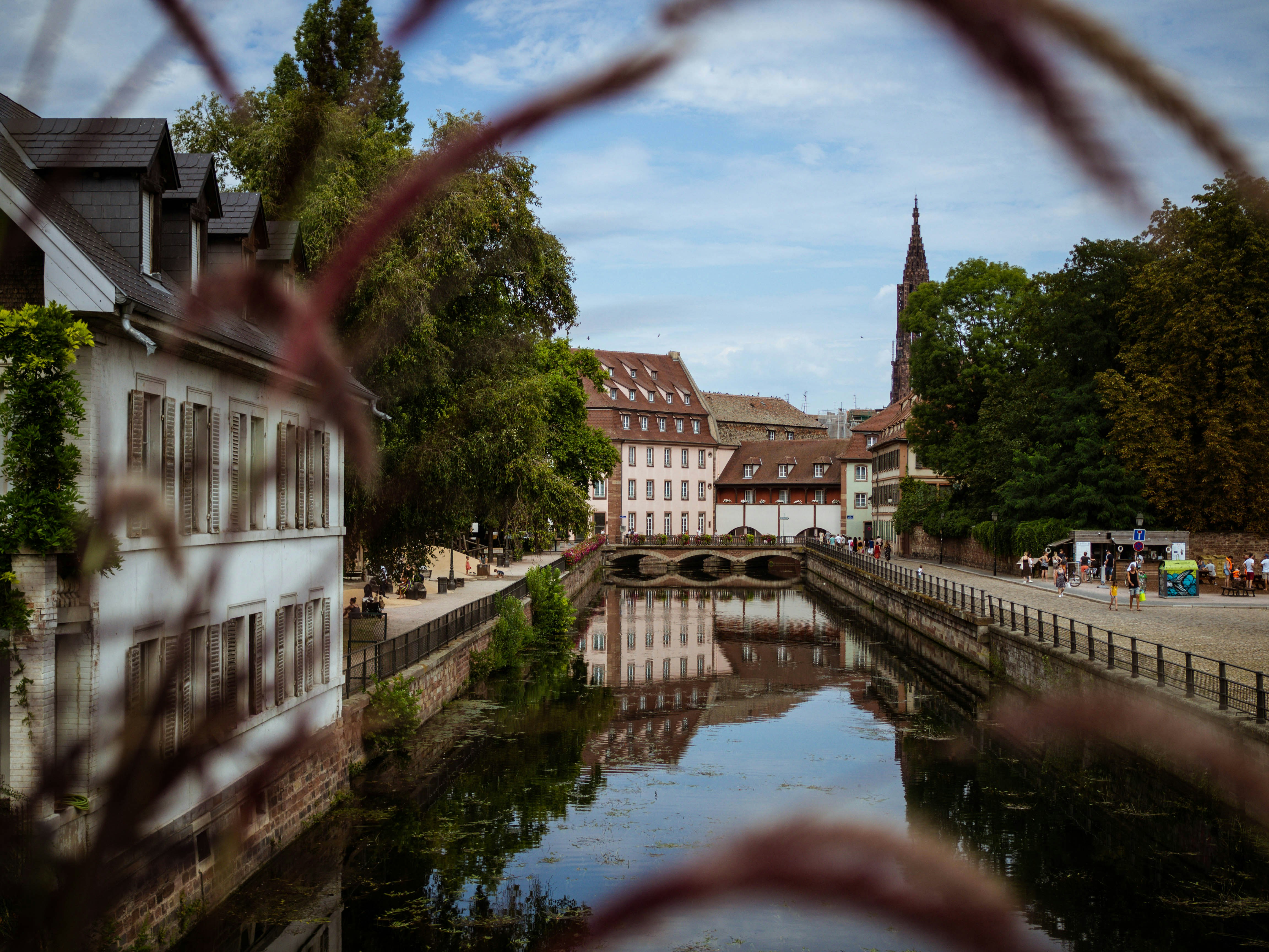 A tranquil canal scene framed by lush greenery, showcasing charming architecture and a bridge reflecting in the water. Visitors enjoy the serene atmosphere along the bank.