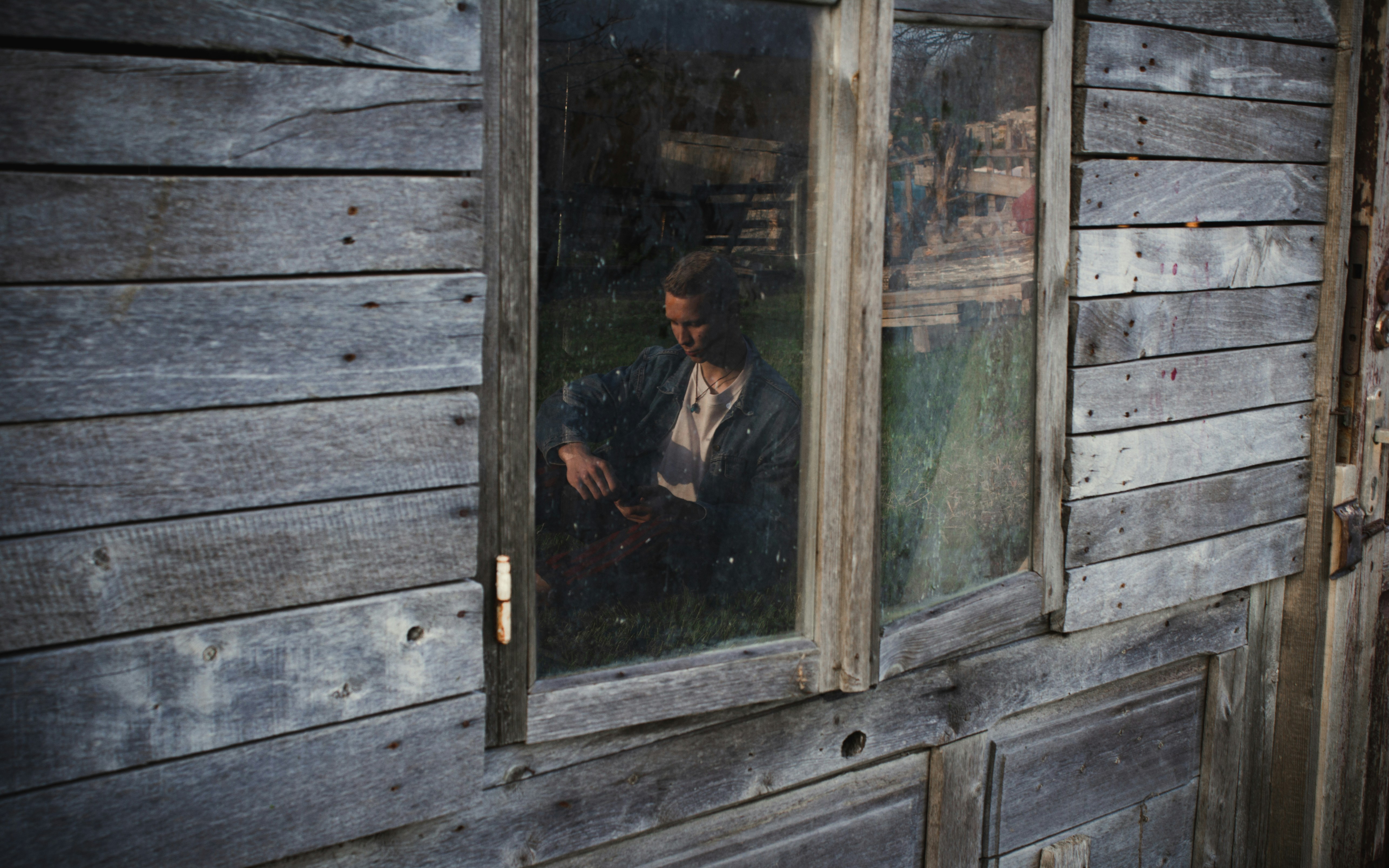 A young person sits contemplatively by a rustic wooden window, their reflection merging with the weathered exterior. The scene evokes a sense of introspection.