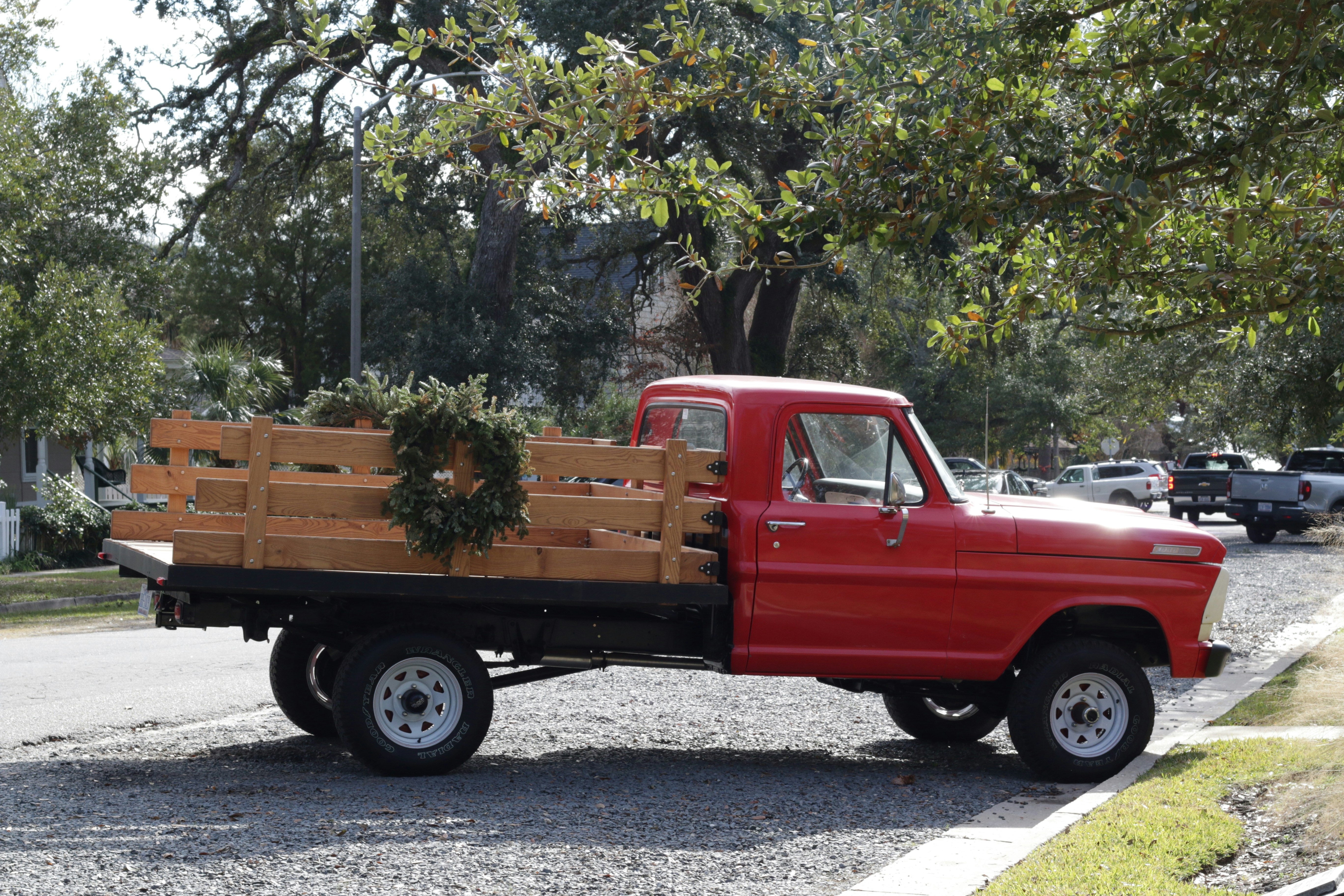 Classic red pickup truck with a wooden flatbed adorned with greenery parked along a tree-lined street.