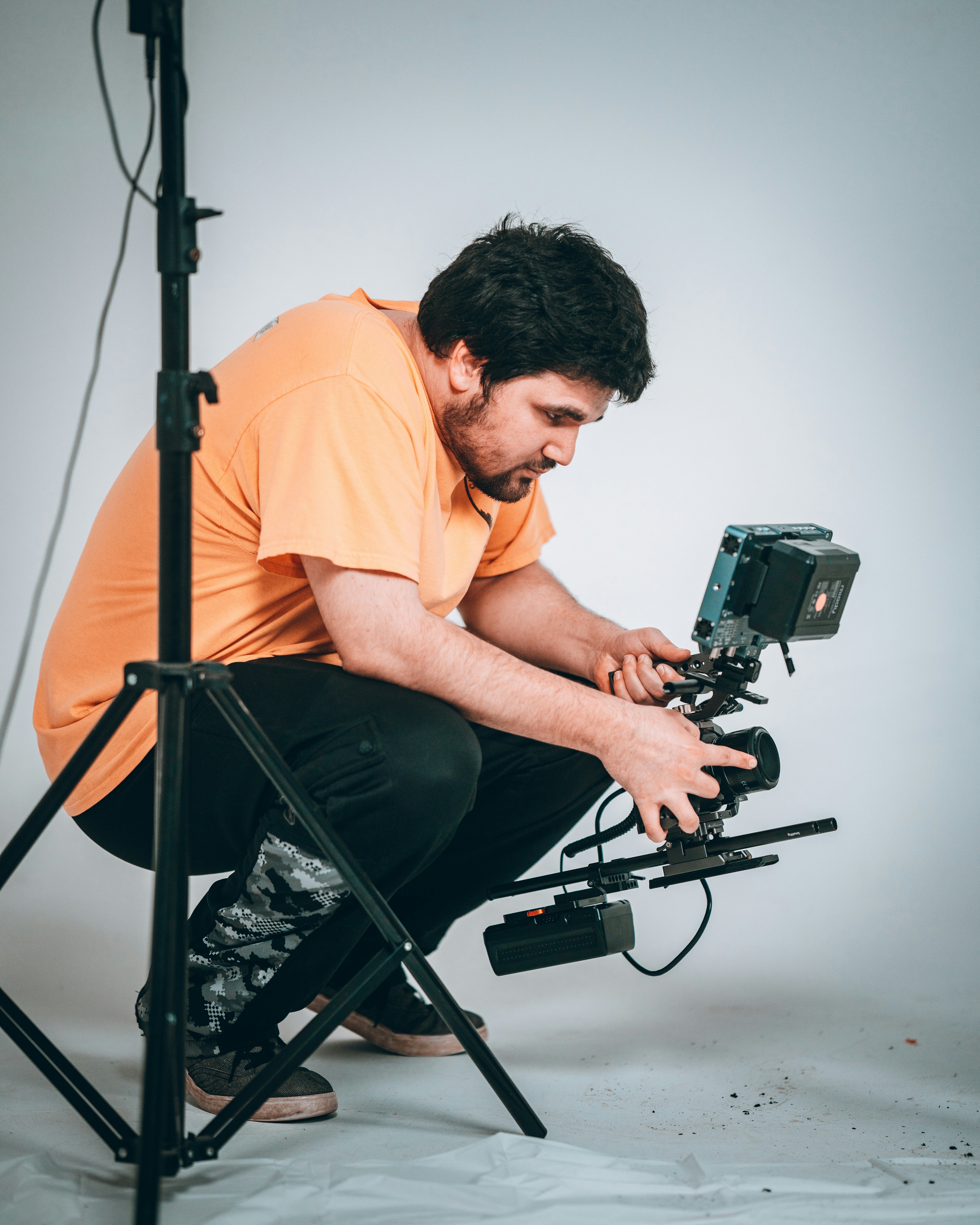 A cinematographer crouches while adjusting a camera rig, focused on achieving the perfect shot. The backdrop is a simple studio setting.