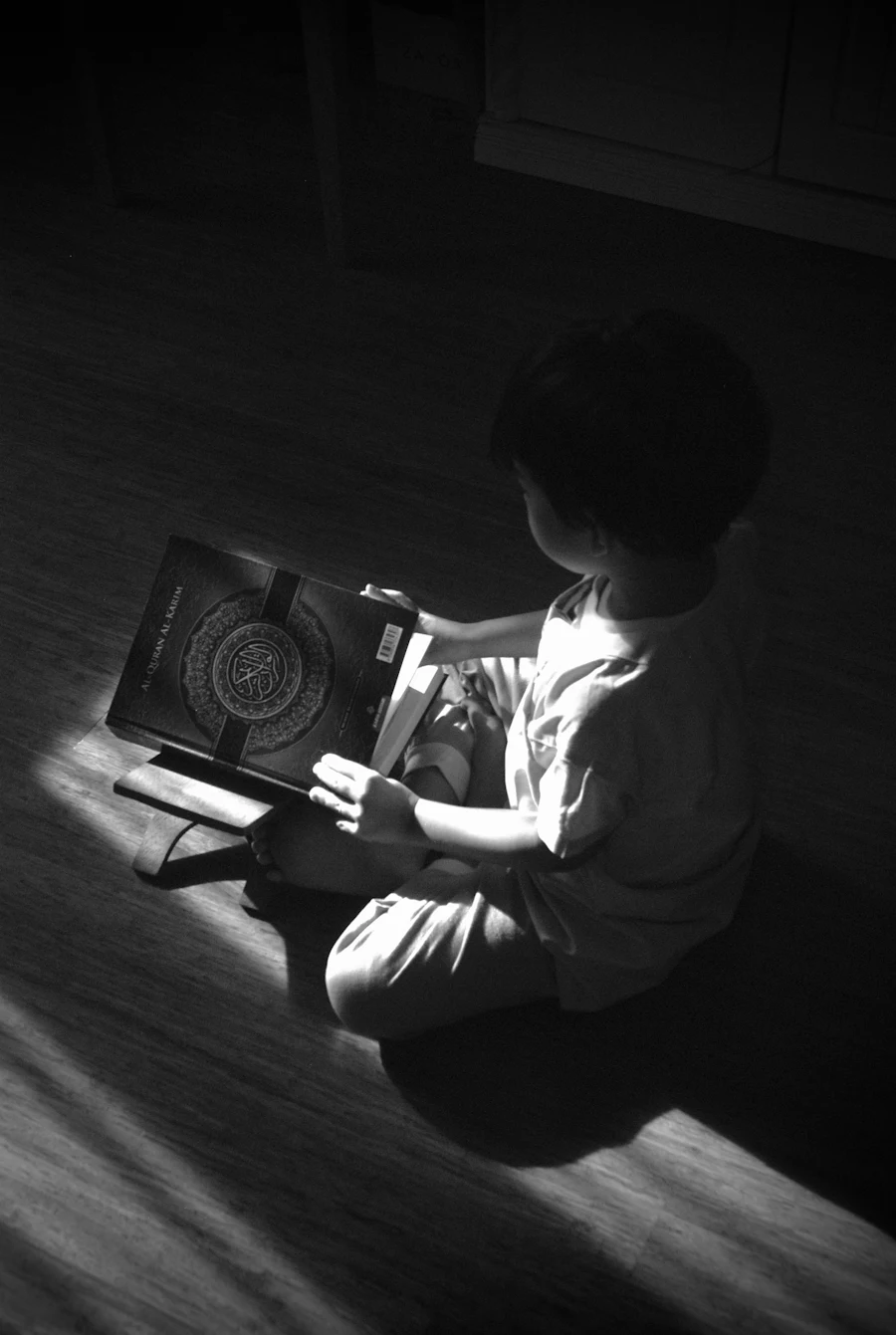 A small child sits on the floor reading a book in a narrow shaft of sunlight, surrounded by deep shadow. The scene is rendered in high-contrast black and white.