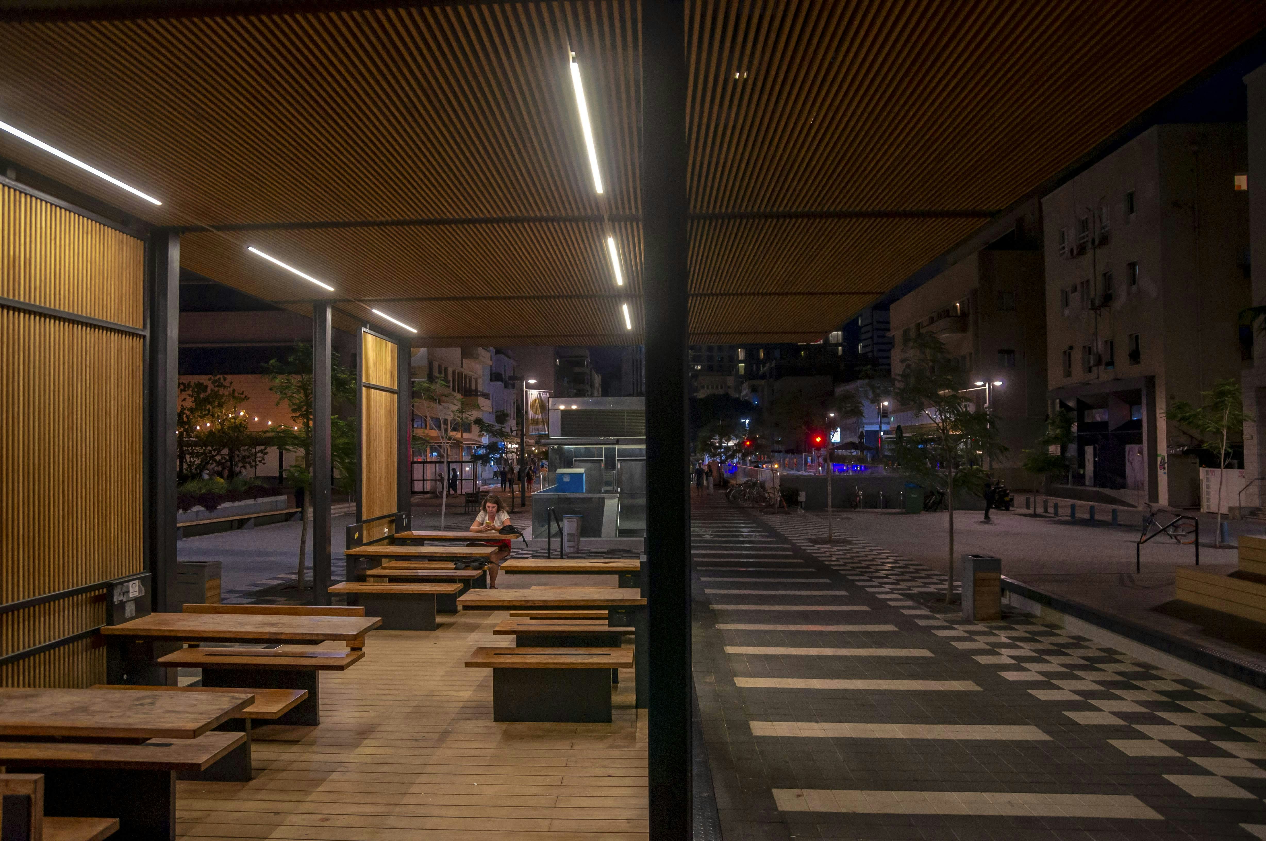 Night scene photograph of a covered wooden terrace with benches and linear lighting, extending toward a crosswalk and distant city street.