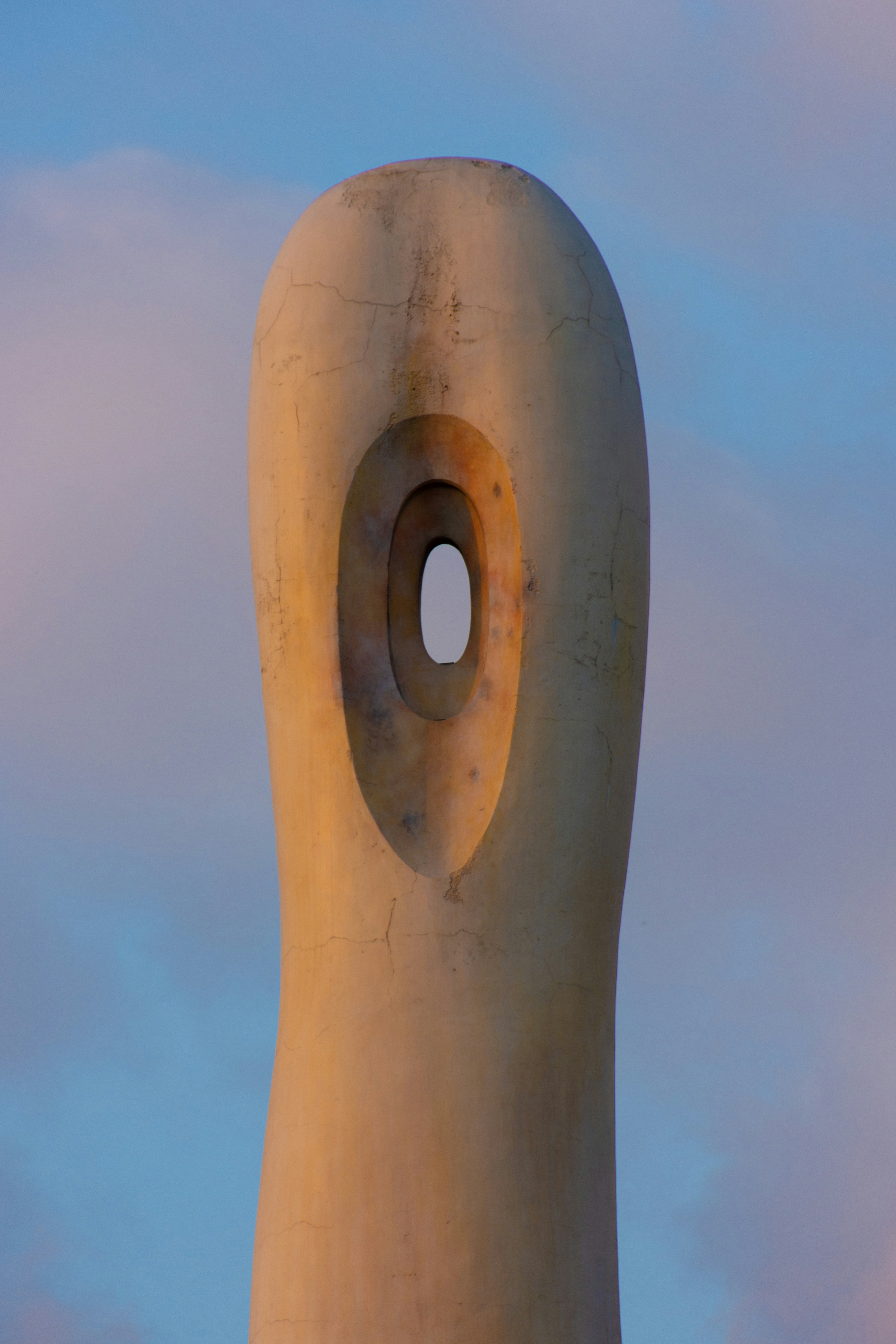 Abstract sculpture resembling a tall tower with an oval opening, set against a pastel sky. The piece invites contemplation on form and perception.