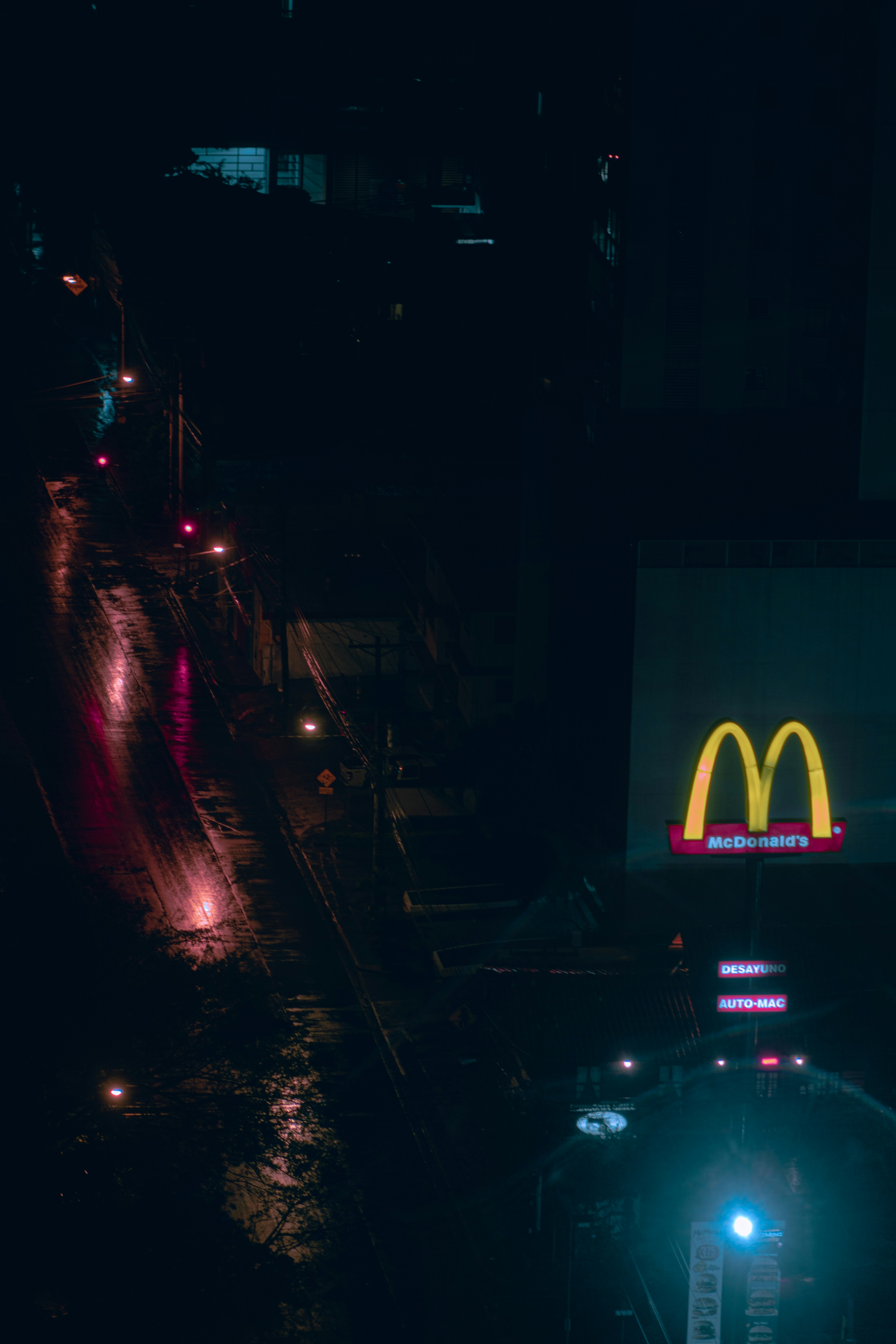 Illuminated McDonald's sign reflecting on a rain-soaked street at night, surrounded by urban lights and shadows.