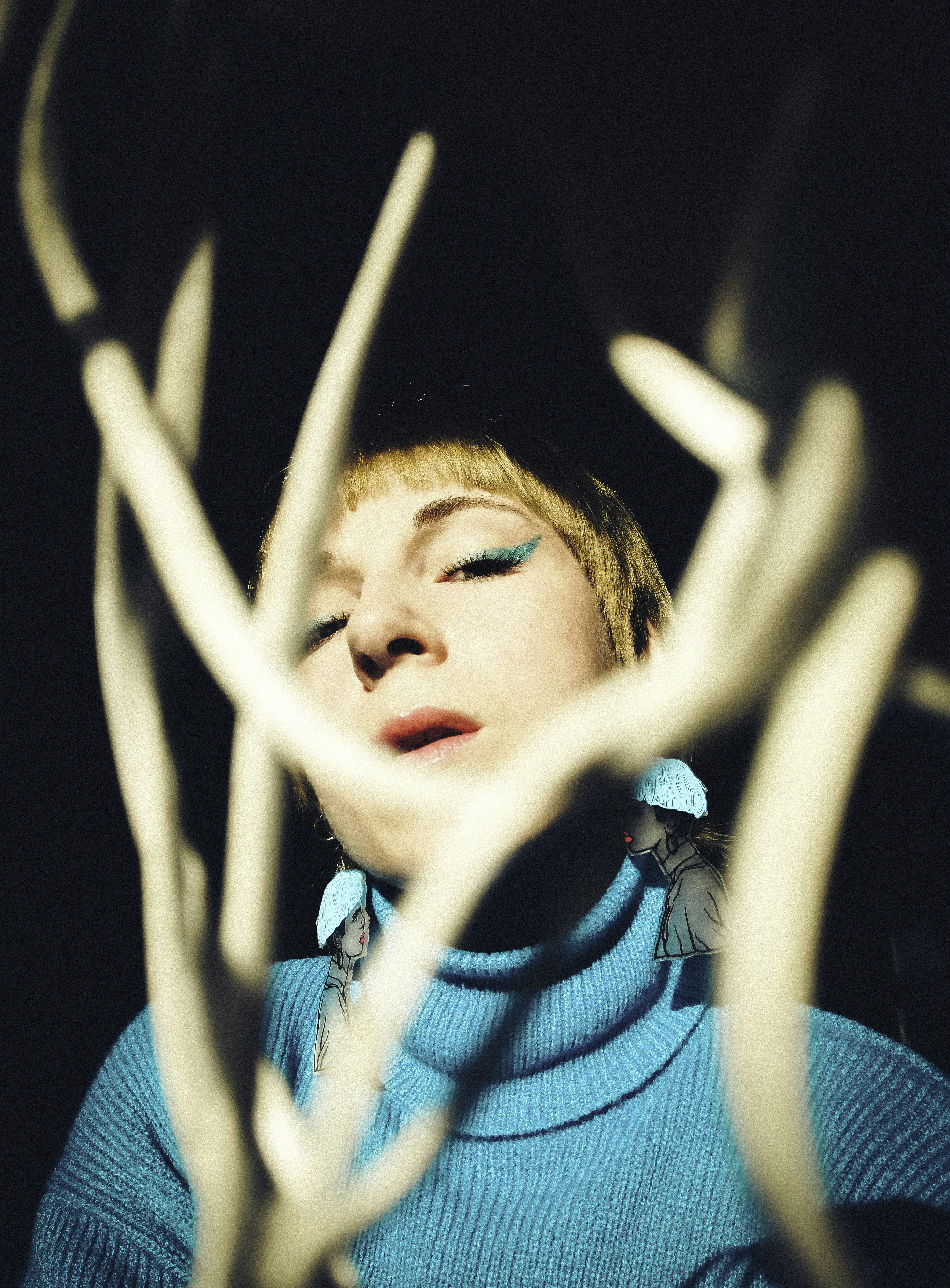 Close-up photograph of a person in a blue turtleneck, seen through pale, out-of-focus branches. The image emphasizes mood and texture against a dark backdrop.