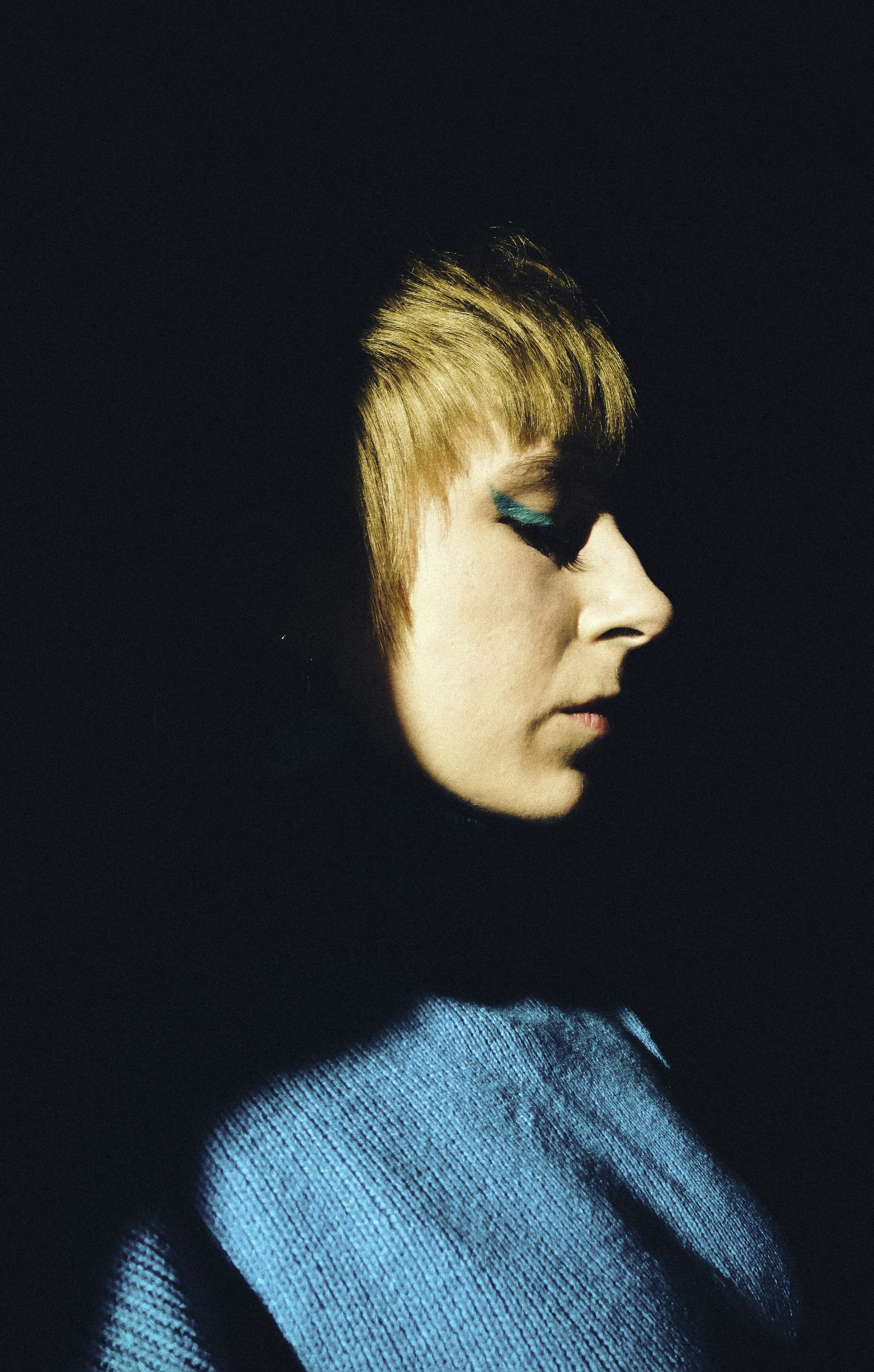 Profile of a woman with striking blue eye makeup, illuminated by a soft light against a dark background.