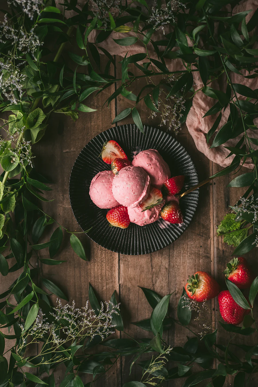 Scoops of pink strawberry ice cream on a black plate, surrounded by fresh strawberries and leafy greenery on a wooden surface.