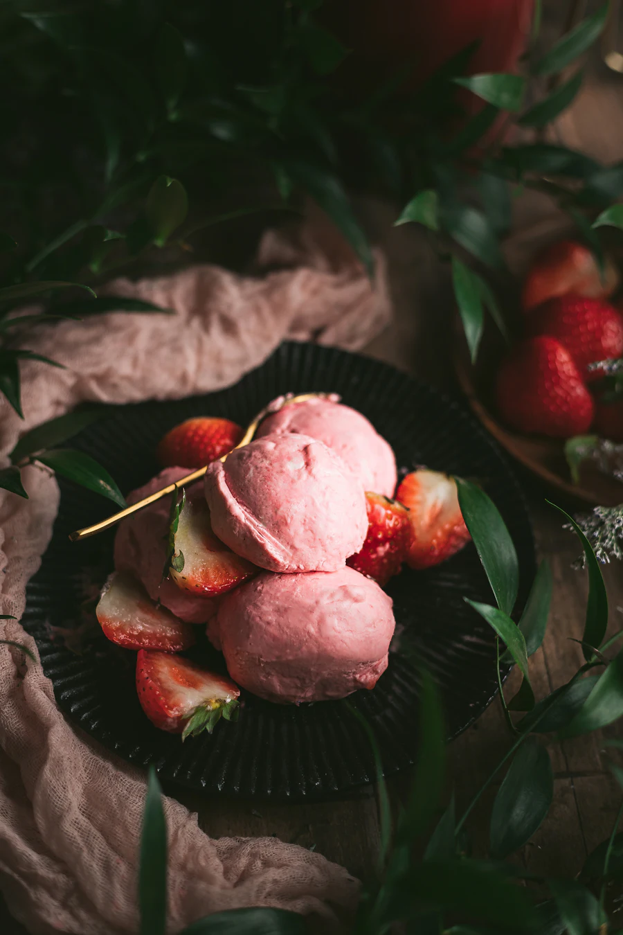 Three scoops of pink strawberry ice cream piled with sliced strawberries on a black plate, framed by soft green leaves and a blush cloth.