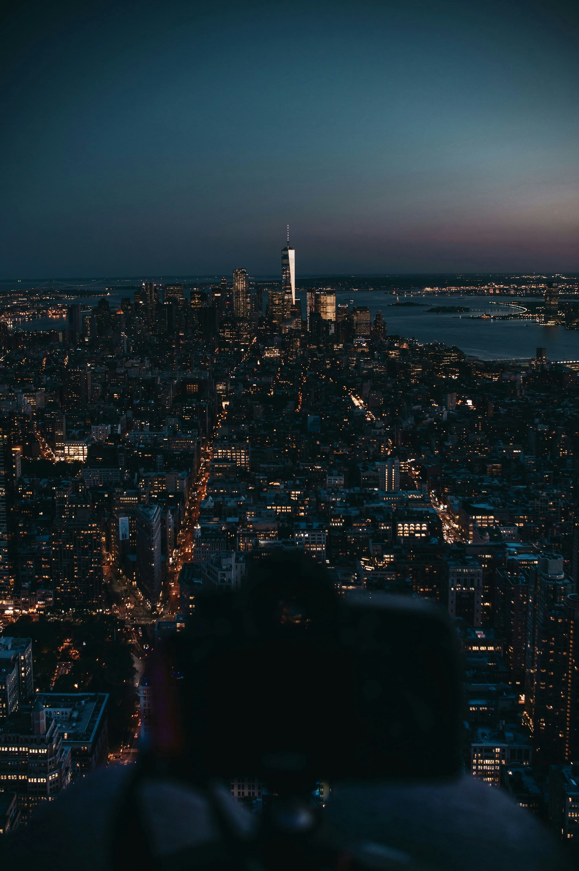 Aerial view of a vibrant city skyline at dusk, with illuminated streets leading to a prominent skyscraper. The scene captures the transition from day to night.