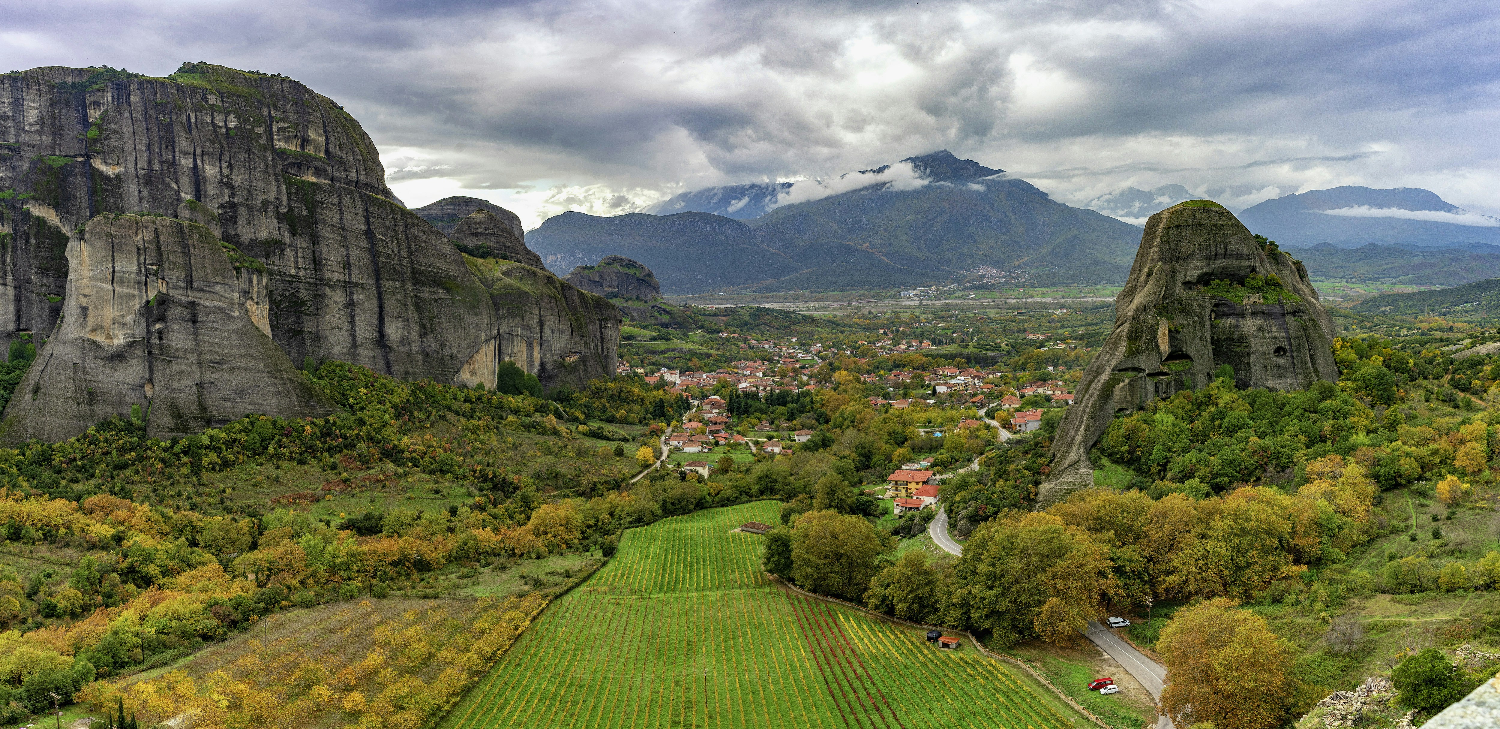 Expansive landscape of Meteora with towering rock formations and lush green fields under a cloudy sky.