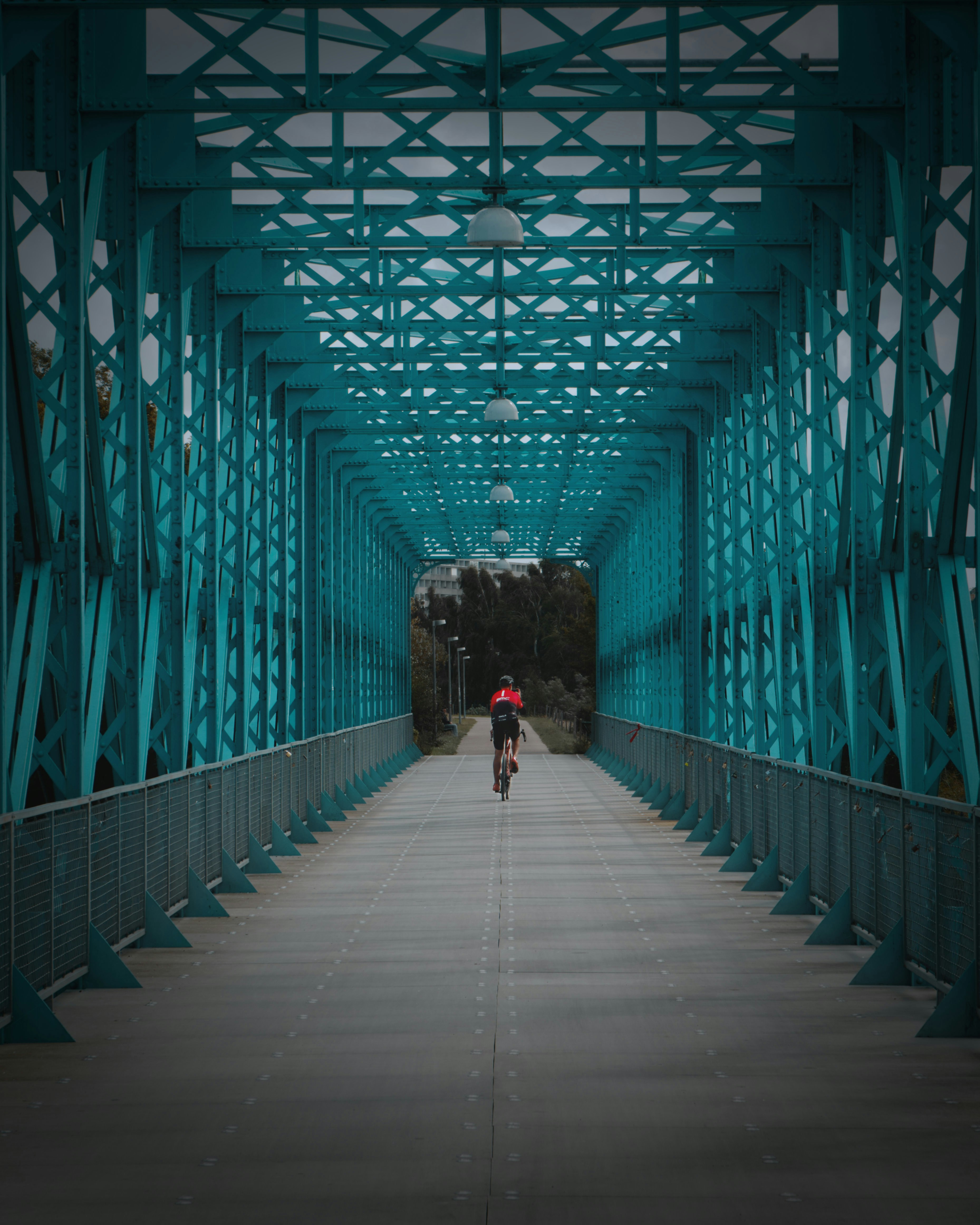 A cyclist rides along a turquoise bridge, surrounded by intricate metalwork and soft lighting. The scene captures the essence of urban exploration and movement.