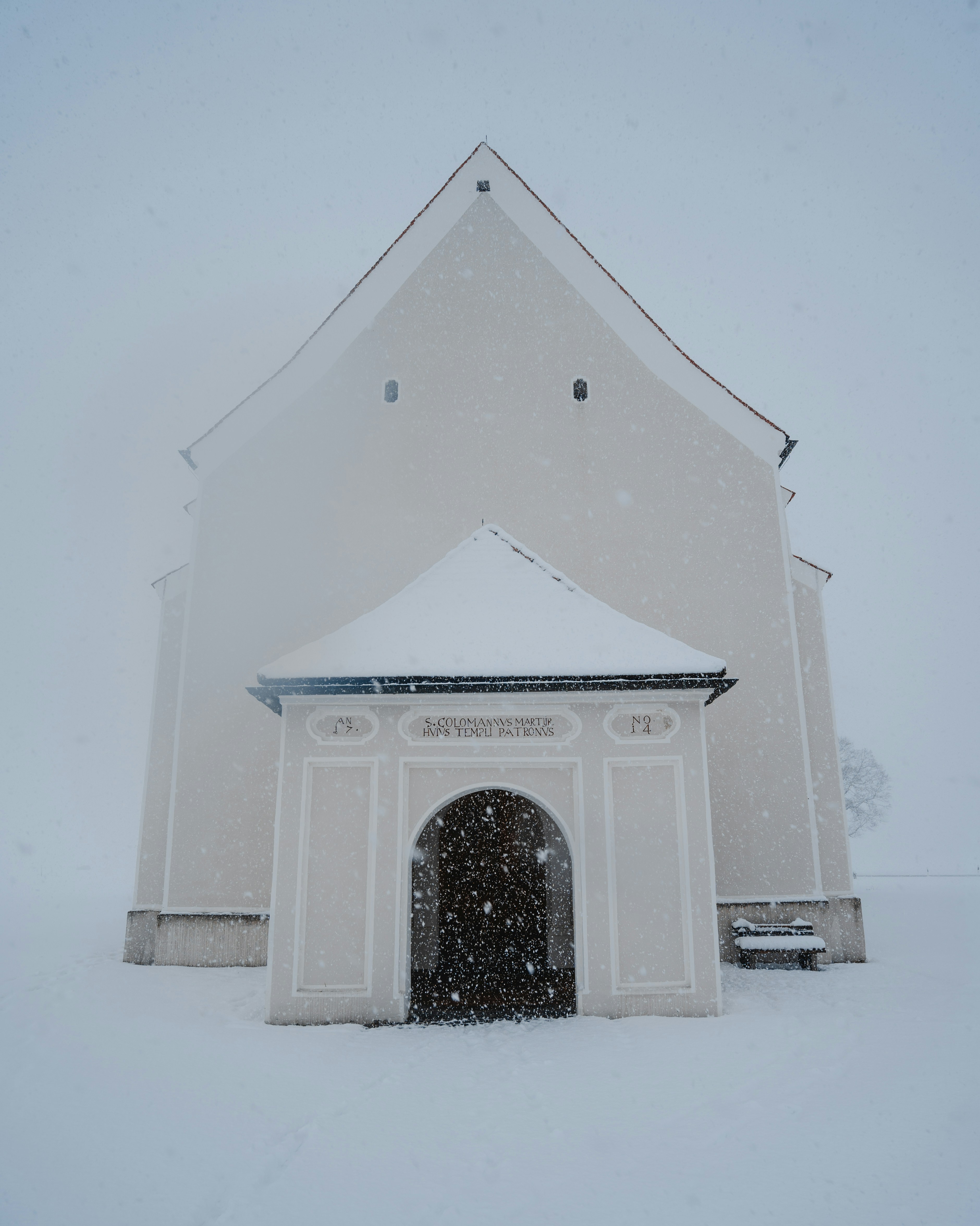 A quaint chapel stands resilient against a heavy snowfall, its entrance framed by swirling snowflakes. The serene winter landscape envelops the structure.