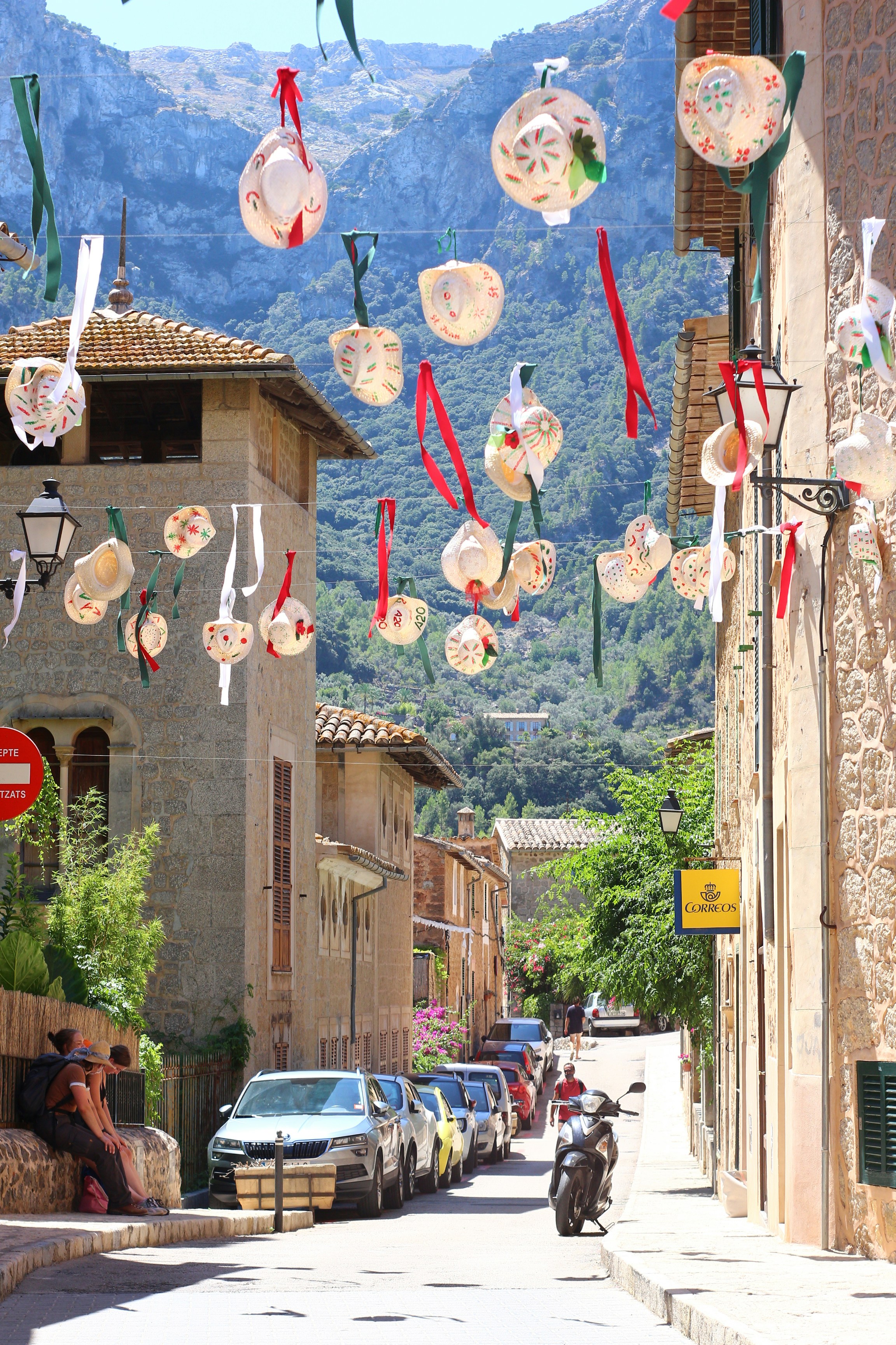 Sunlit street in a European village with decorative paper lanterns hanging above, stone buildings, and parked cars along a narrow lane.