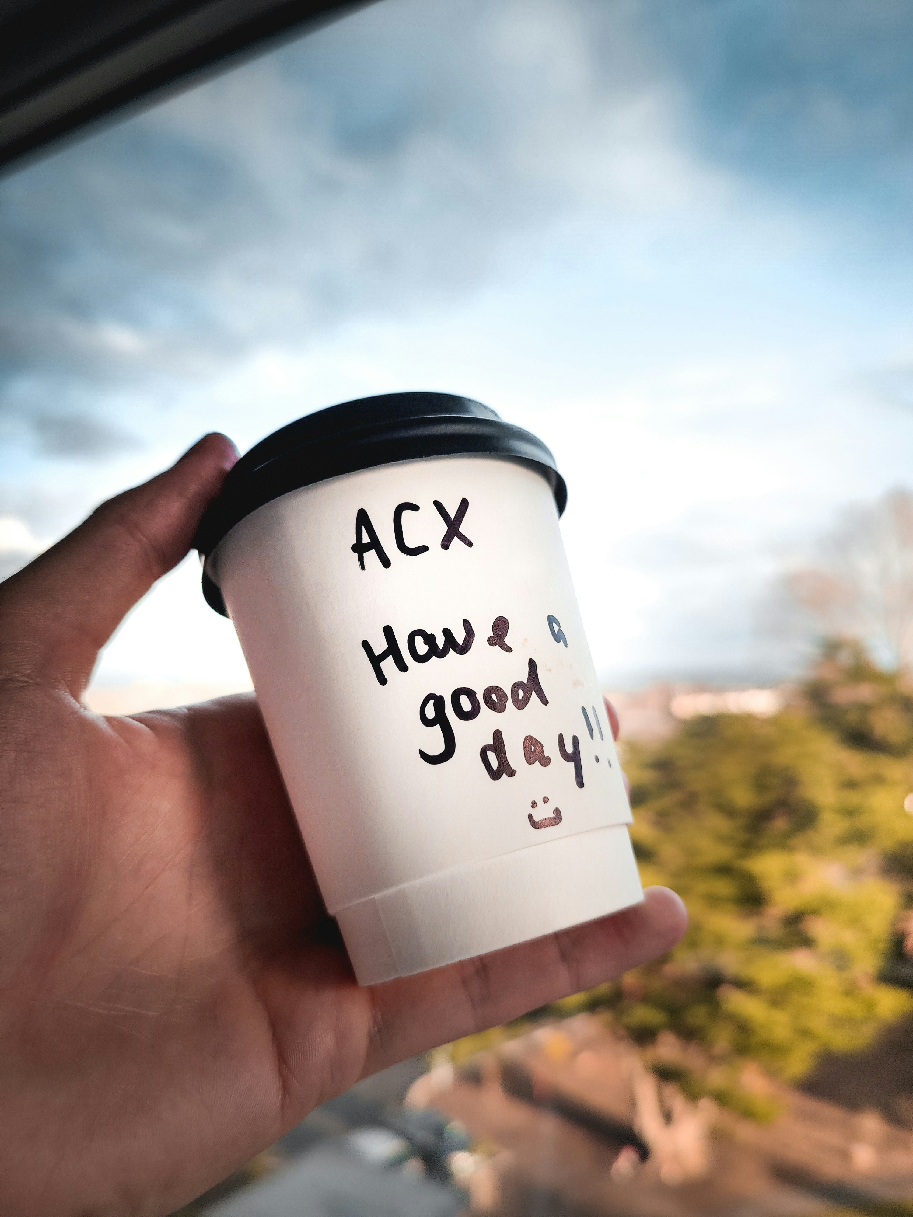 Hand-held disposable coffee cup with handwritten message against a softly blurred outdoor backdrop.