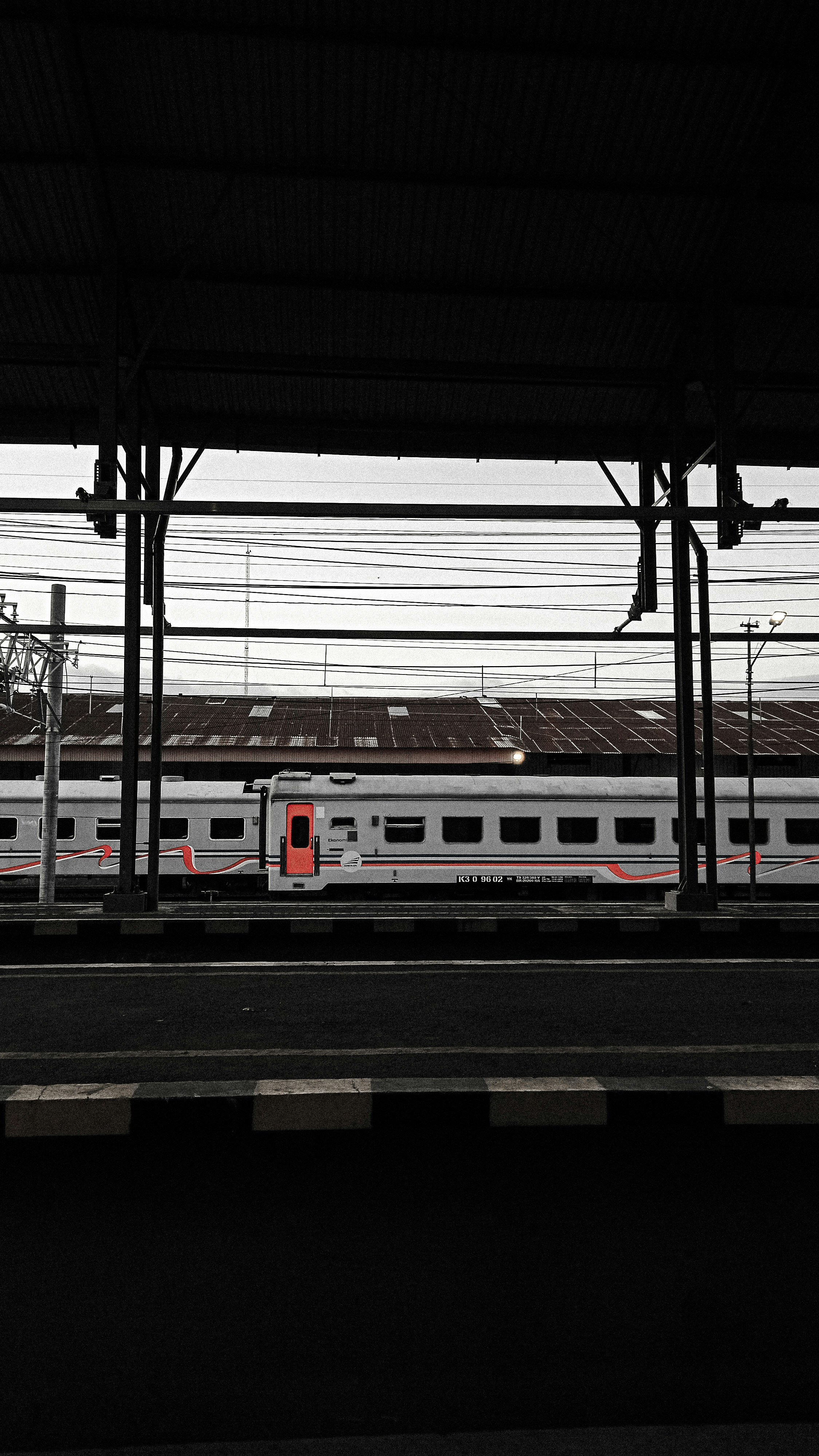 A sleek train stands against a backdrop of industrial architecture, framed by station structures and power lines. The vibrant door adds a striking contrast to the subdued tones.