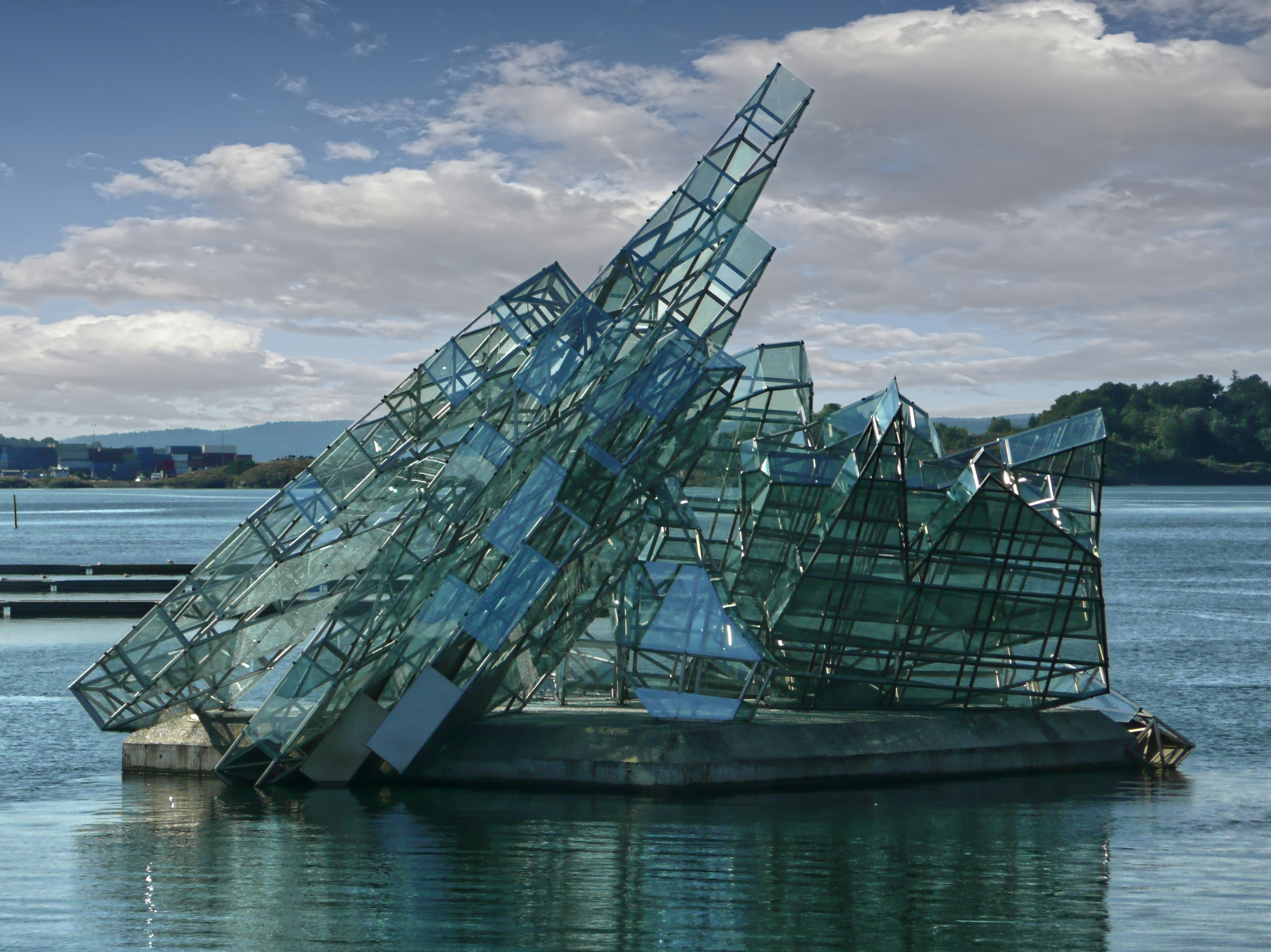 Abstract glass sculpture emerging from tranquil waters, showcasing unique geometric forms against a cloudy sky.
