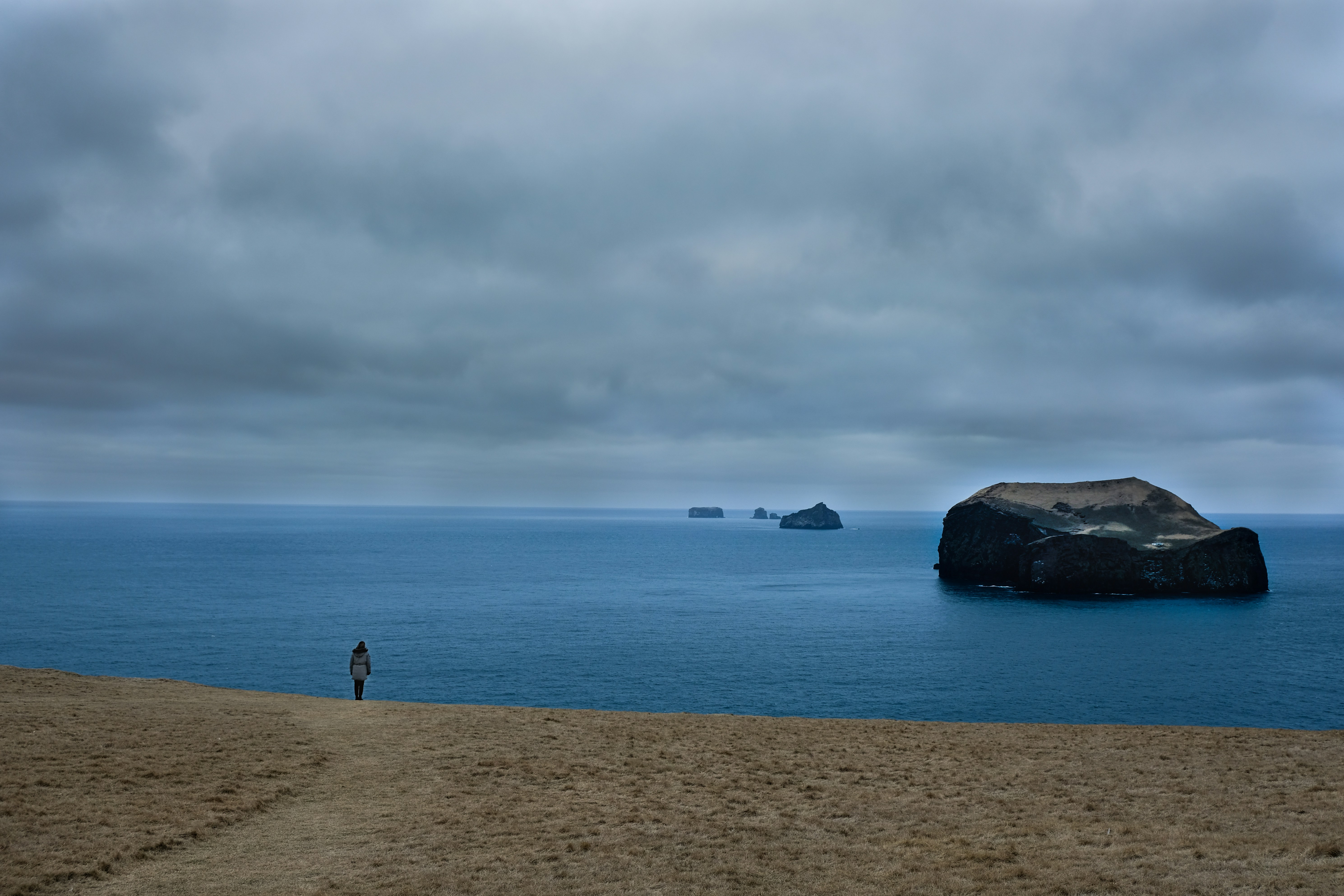Person standing on a cliff overlooking the vast ocean and distant islands under a cloudy sky.