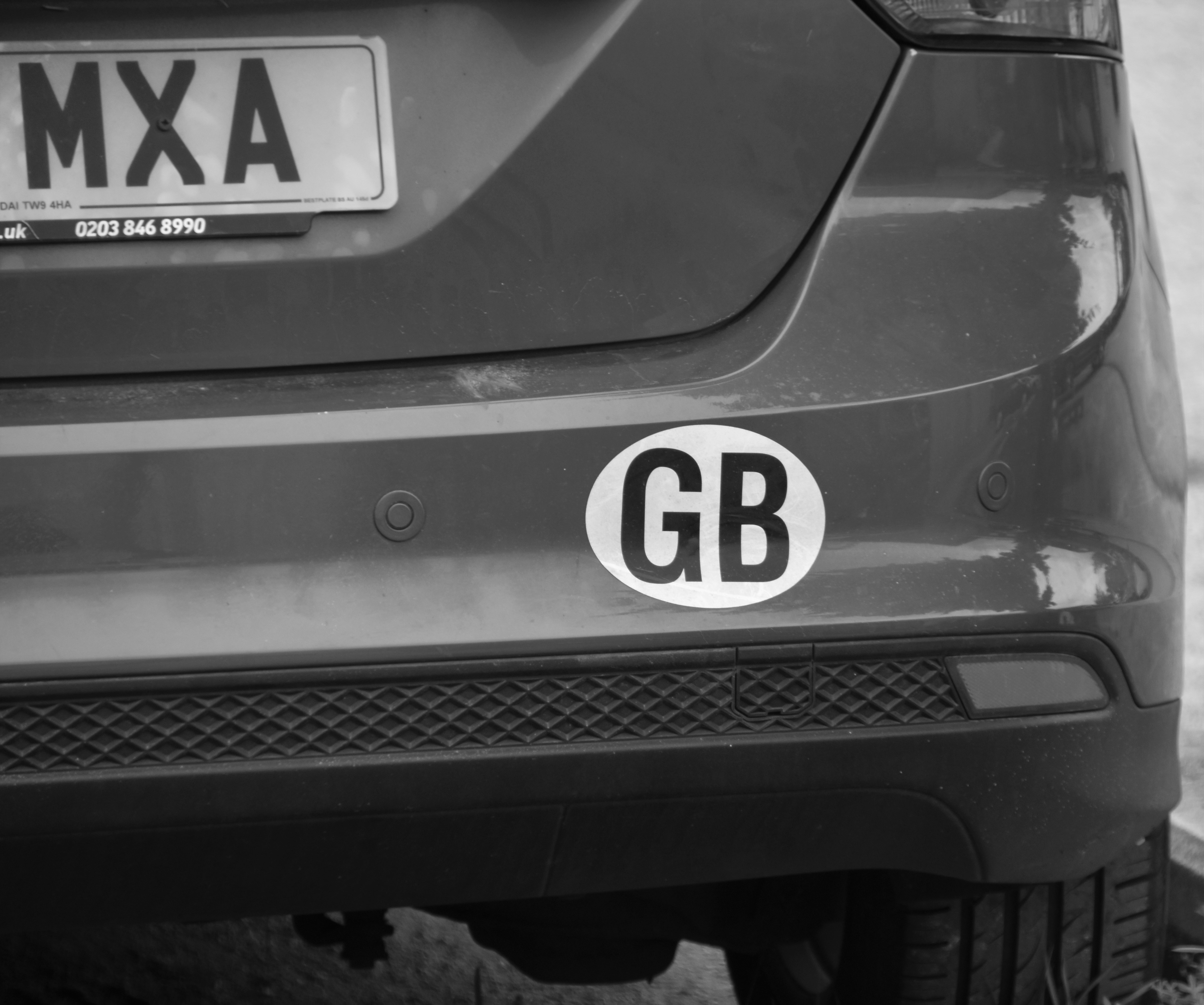 A close-up of a car's rear bumper featuring a circular 'GB' sticker, emphasizing national identity. The monochrome effect adds a classic touch.