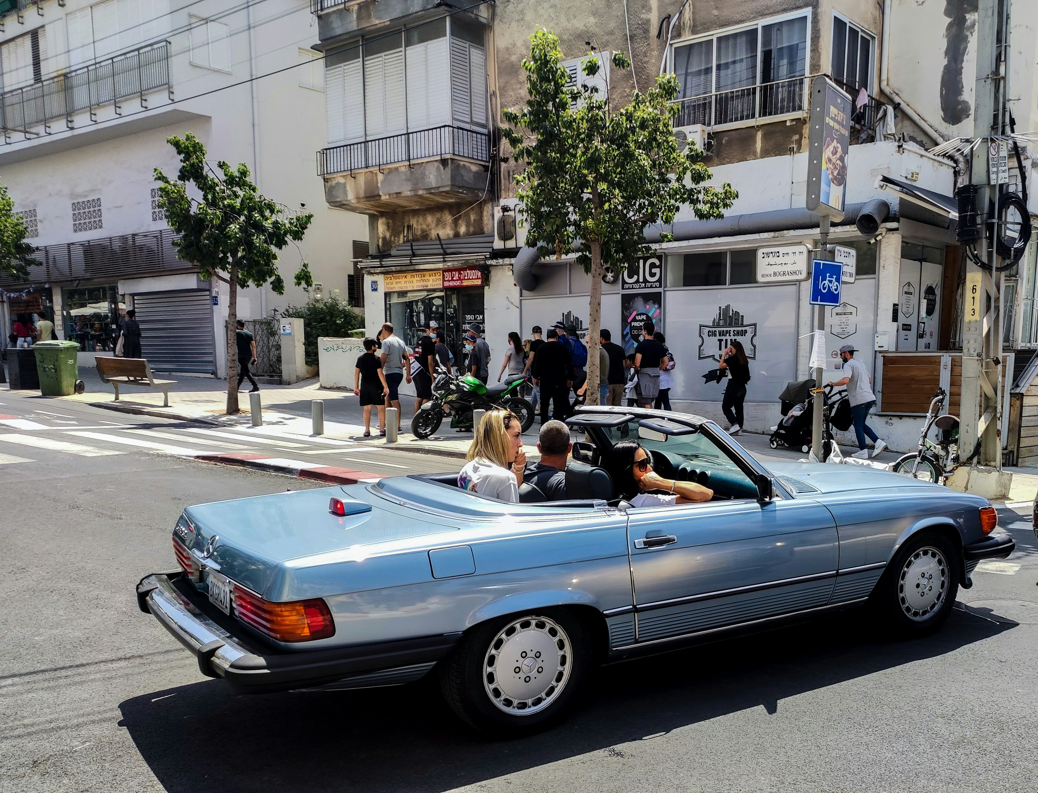 Classic convertible car navigating a lively street scene filled with pedestrians and shops. The sunlit atmosphere enhances the vibrant city life.