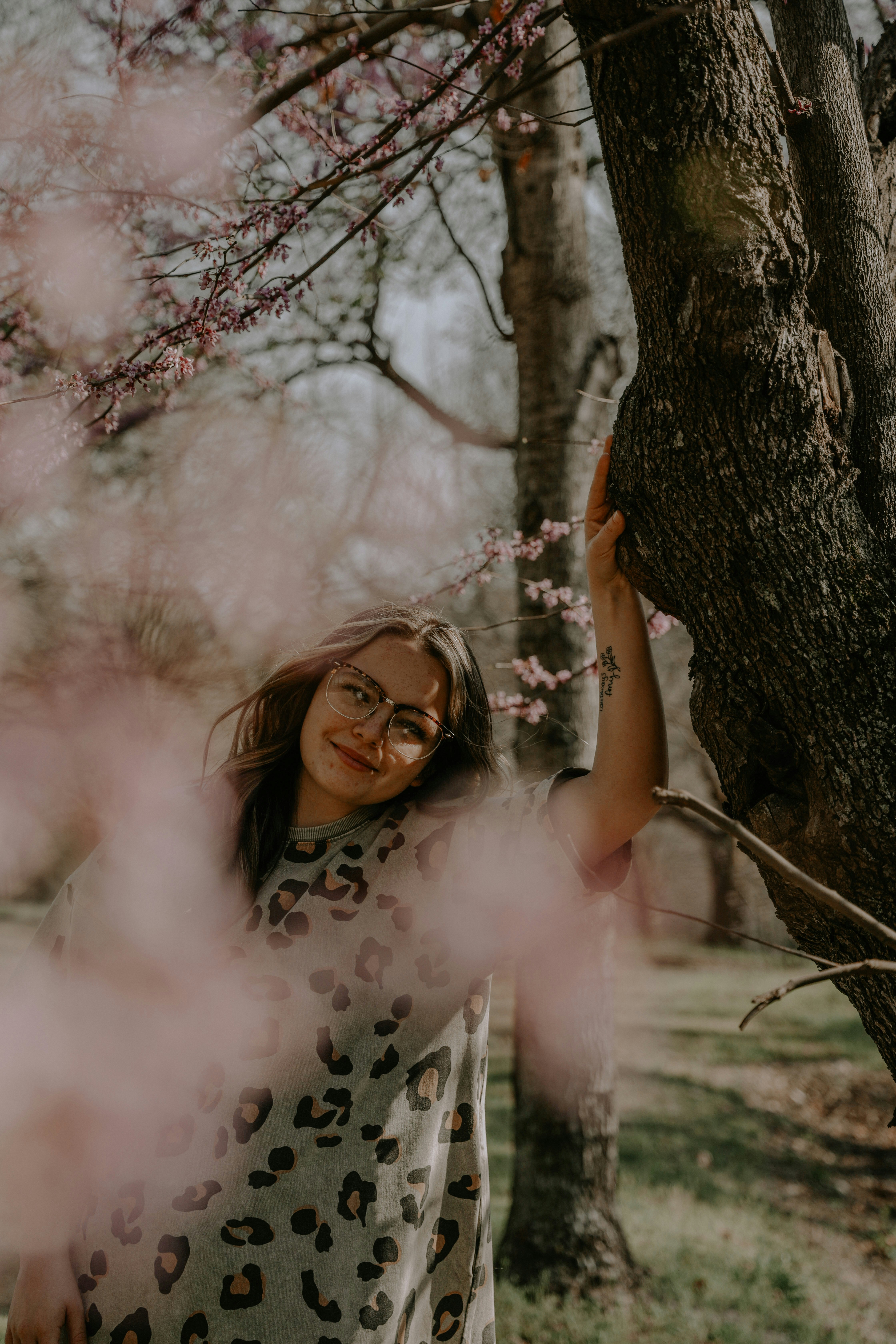 A young woman in a leopard-print outfit stands beside a tree, framed by delicate pink blossoms, exuding a sense of calm and connection to nature.