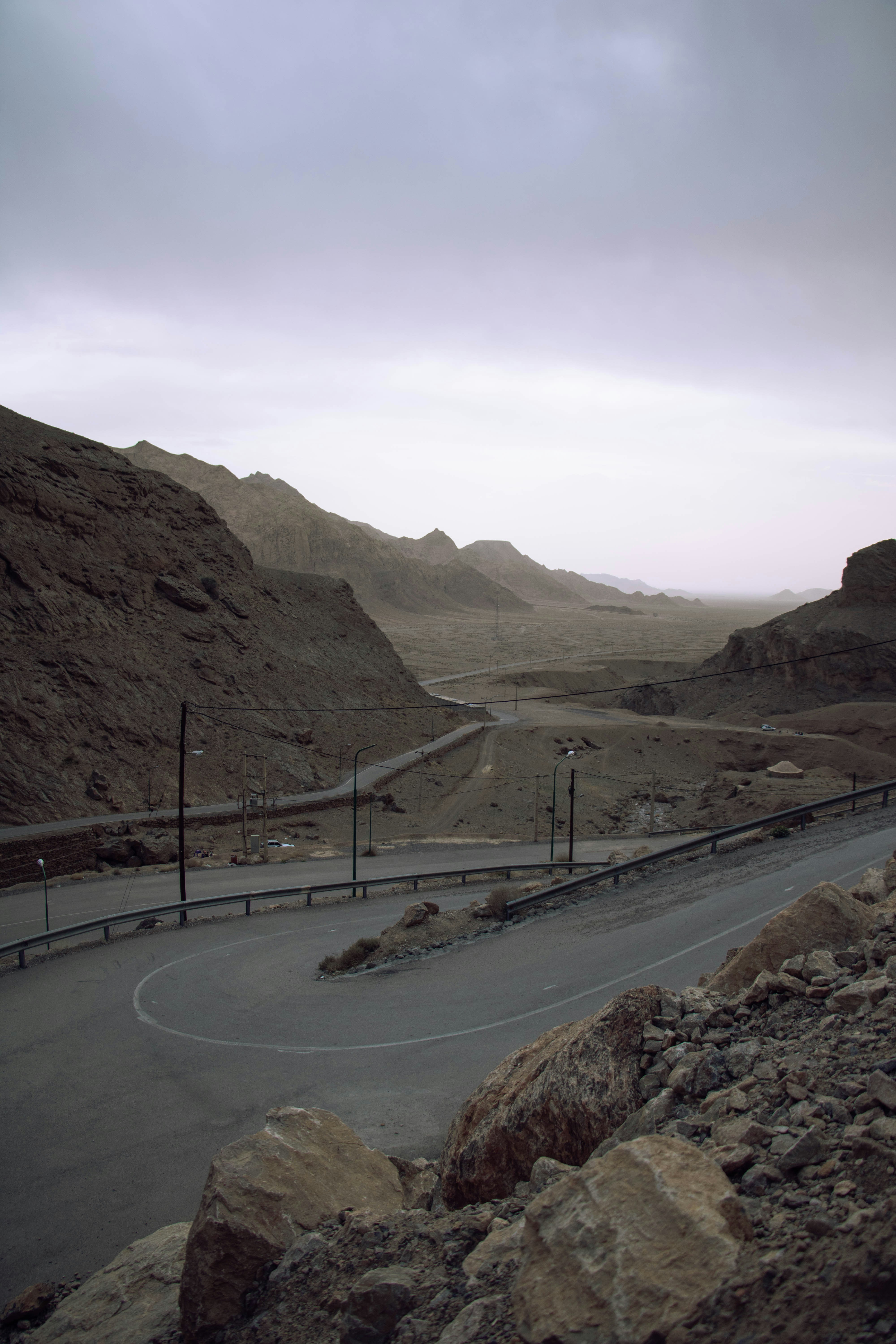 Curved road meandering through rugged terrain, with mountains rising in the background under a cloudy sky.
