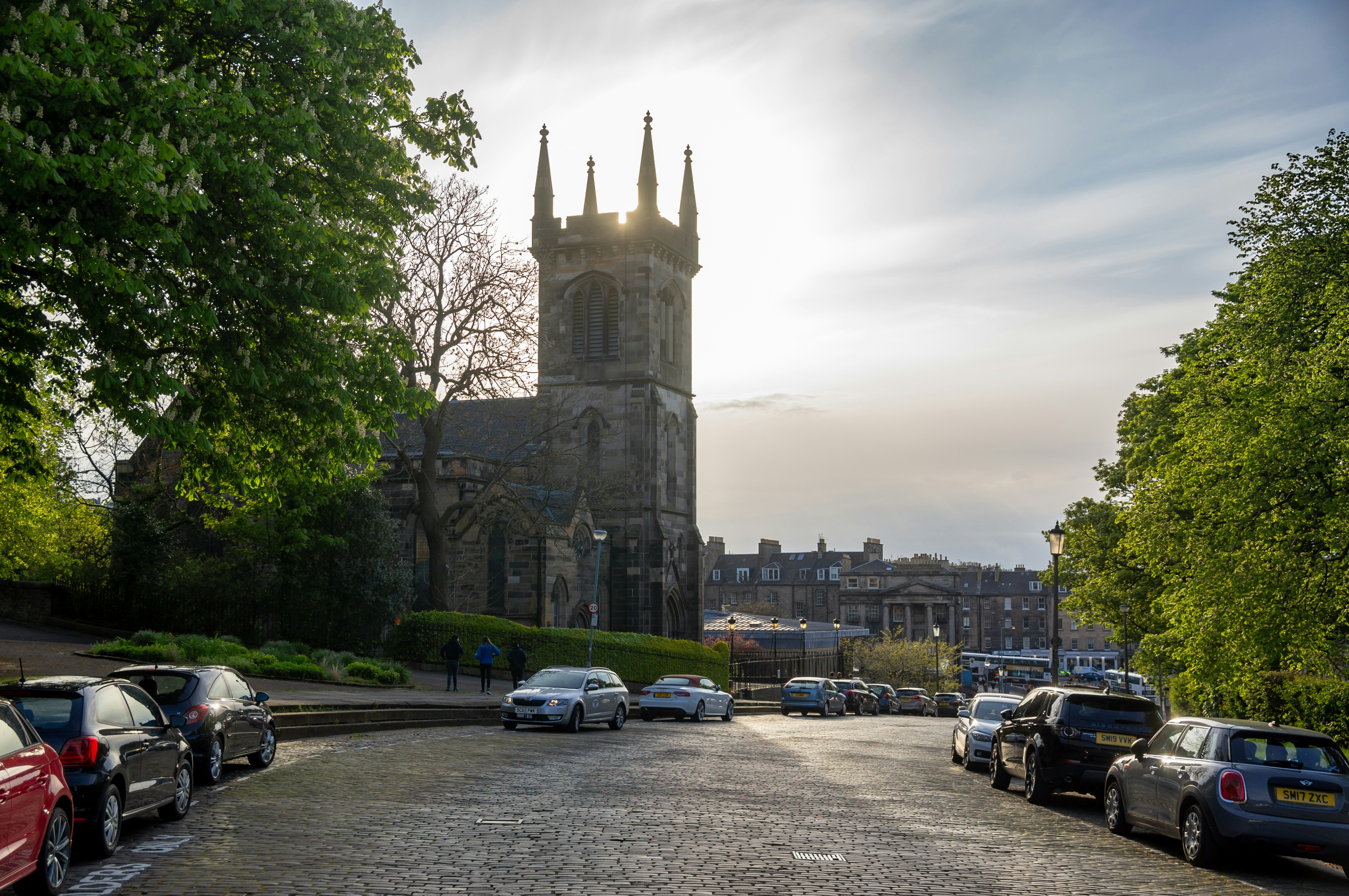 Historic church tower stands prominently against a backdrop of modern architecture, framed by lush greenery and parked cars.