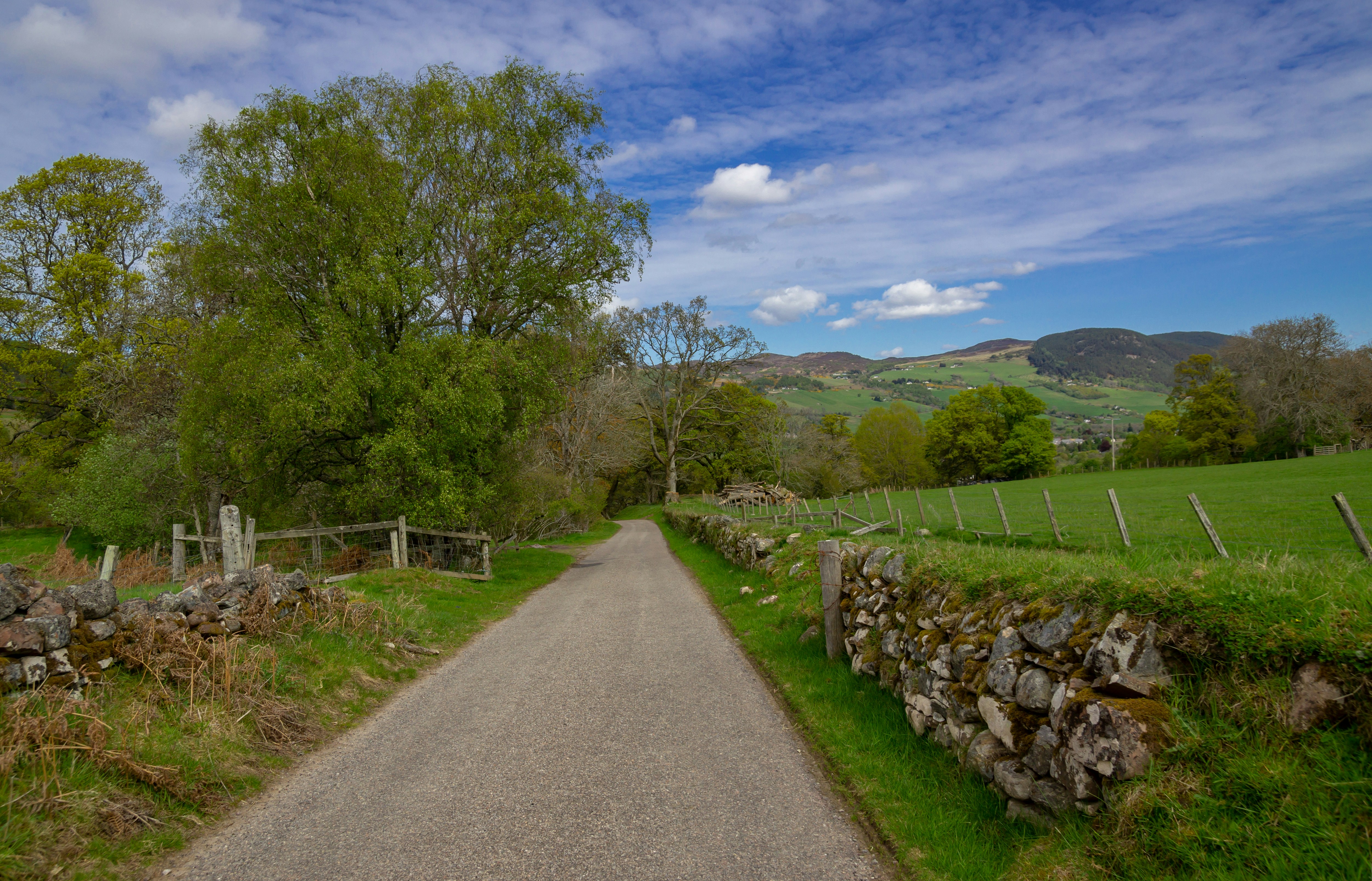 Sunlit country road runs between mossy stone walls, flanked by green fields and trees. A cloud-splashed blue sky crowns the tranquil rural landscape.