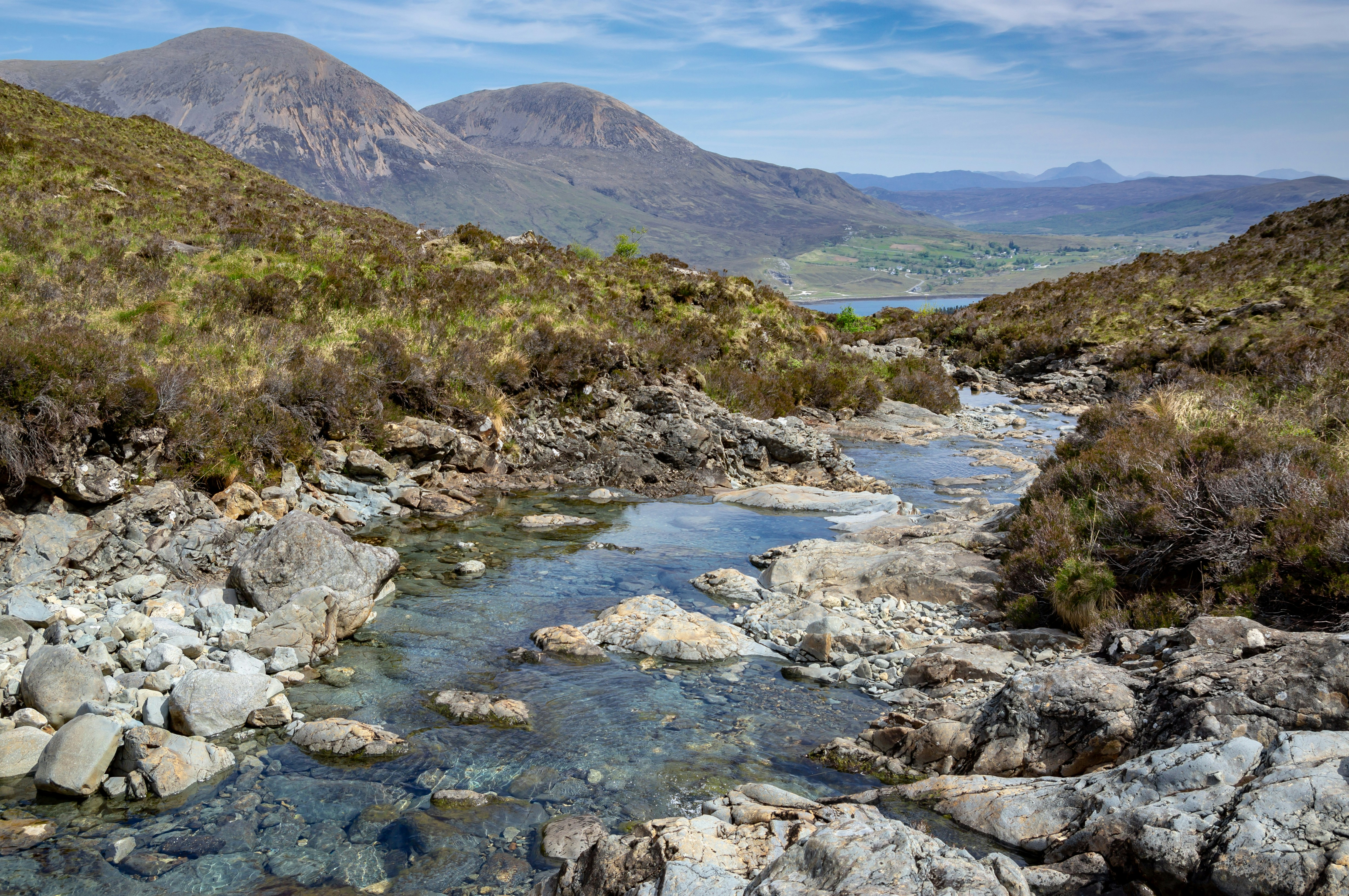 Sunlit mountain stream winds through rocky terrain toward distant hills and a lakeside village, framed by rugged scrub under a bright sky.