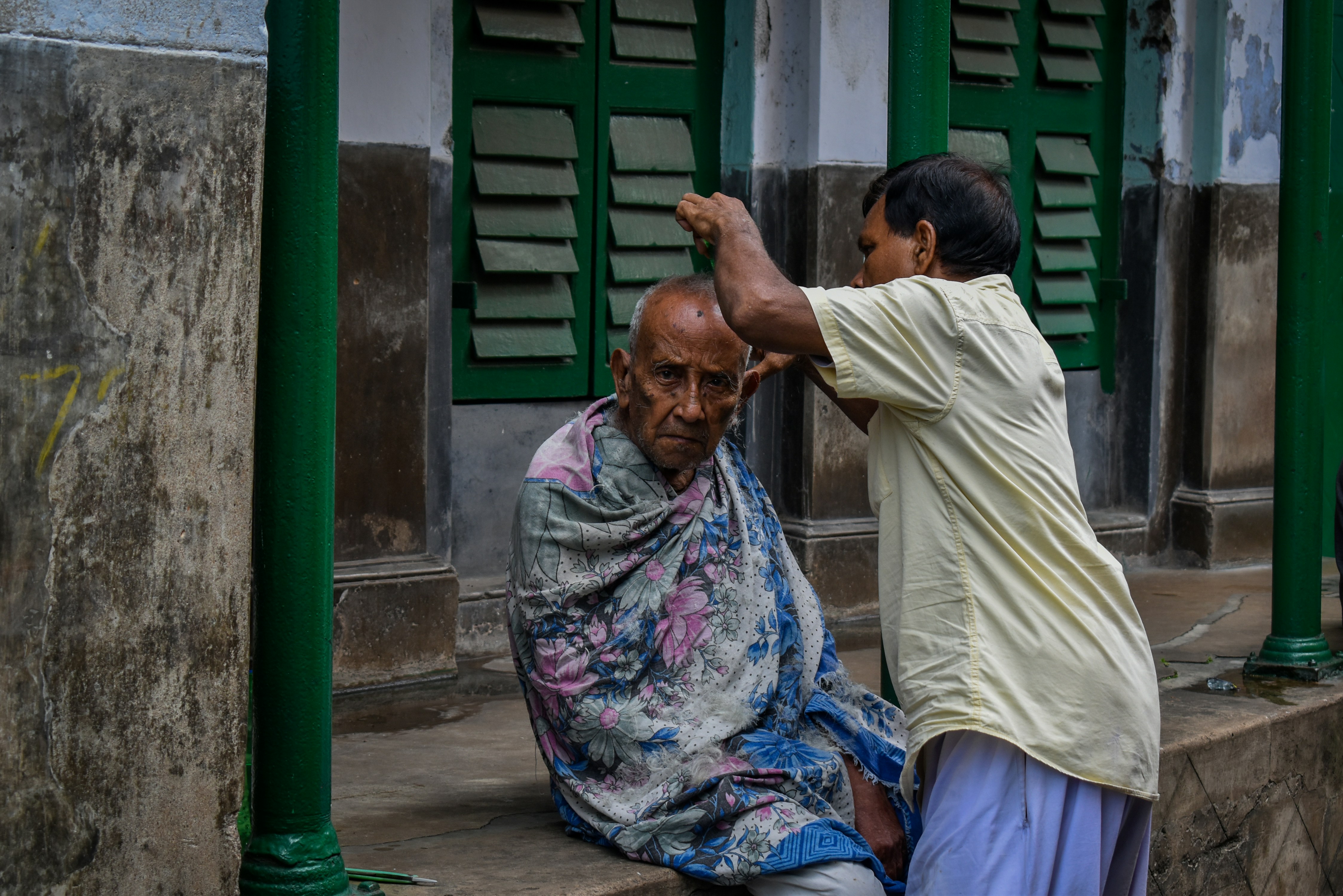 This evocative image captures a candid moment between a barber and his elderly client on a rustic street corner. The composition draws attention to the vibrant contrast of the client's floral shawl against the muted tones of the weathered walls and green shutters. The natural lighting casts soft shadows, adding depth and an intimate atmosphere to this everyday scene, making it visually compelling.