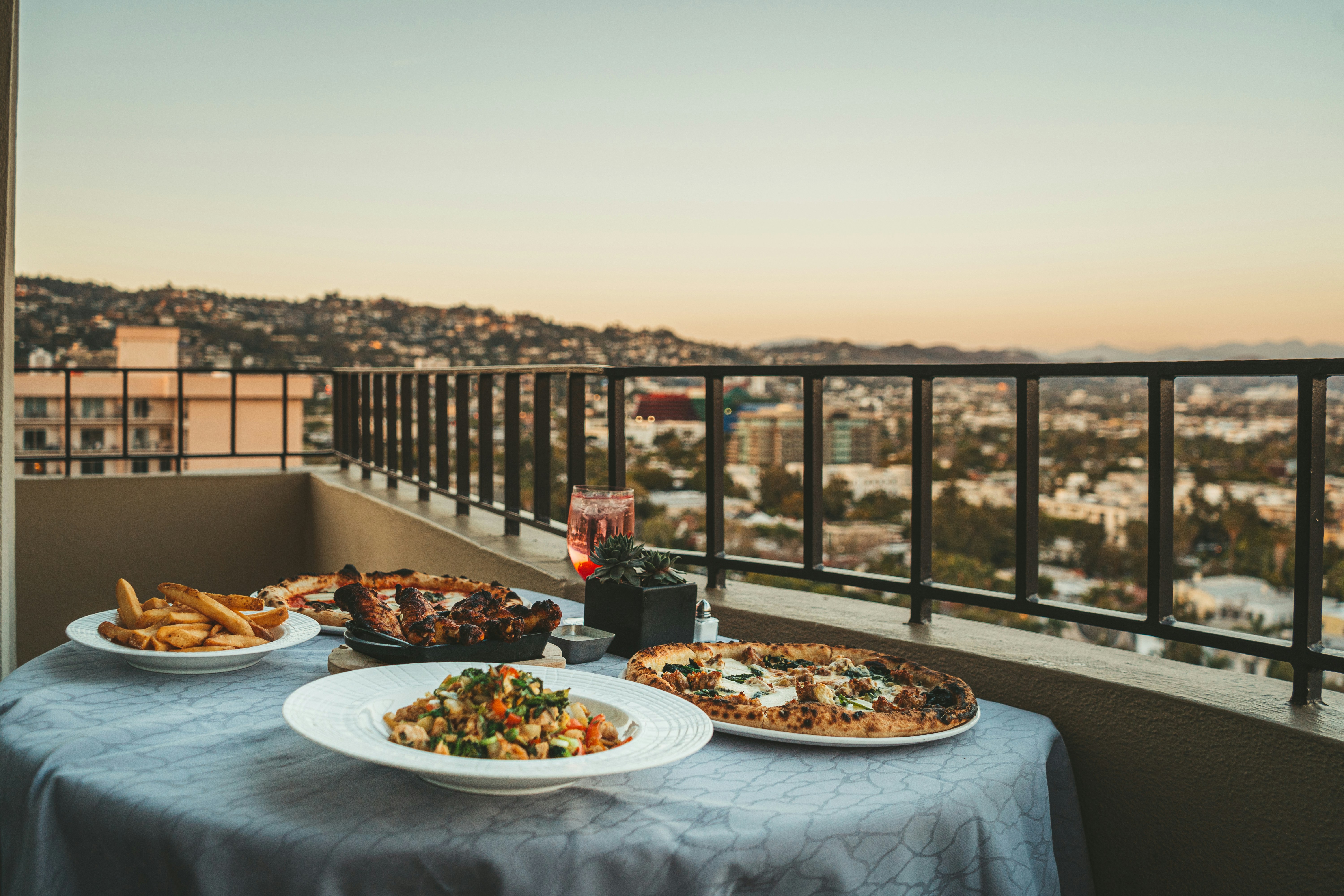 A beautifully arranged table featuring an array of dishes and drinks, set against a panoramic city view at dusk.