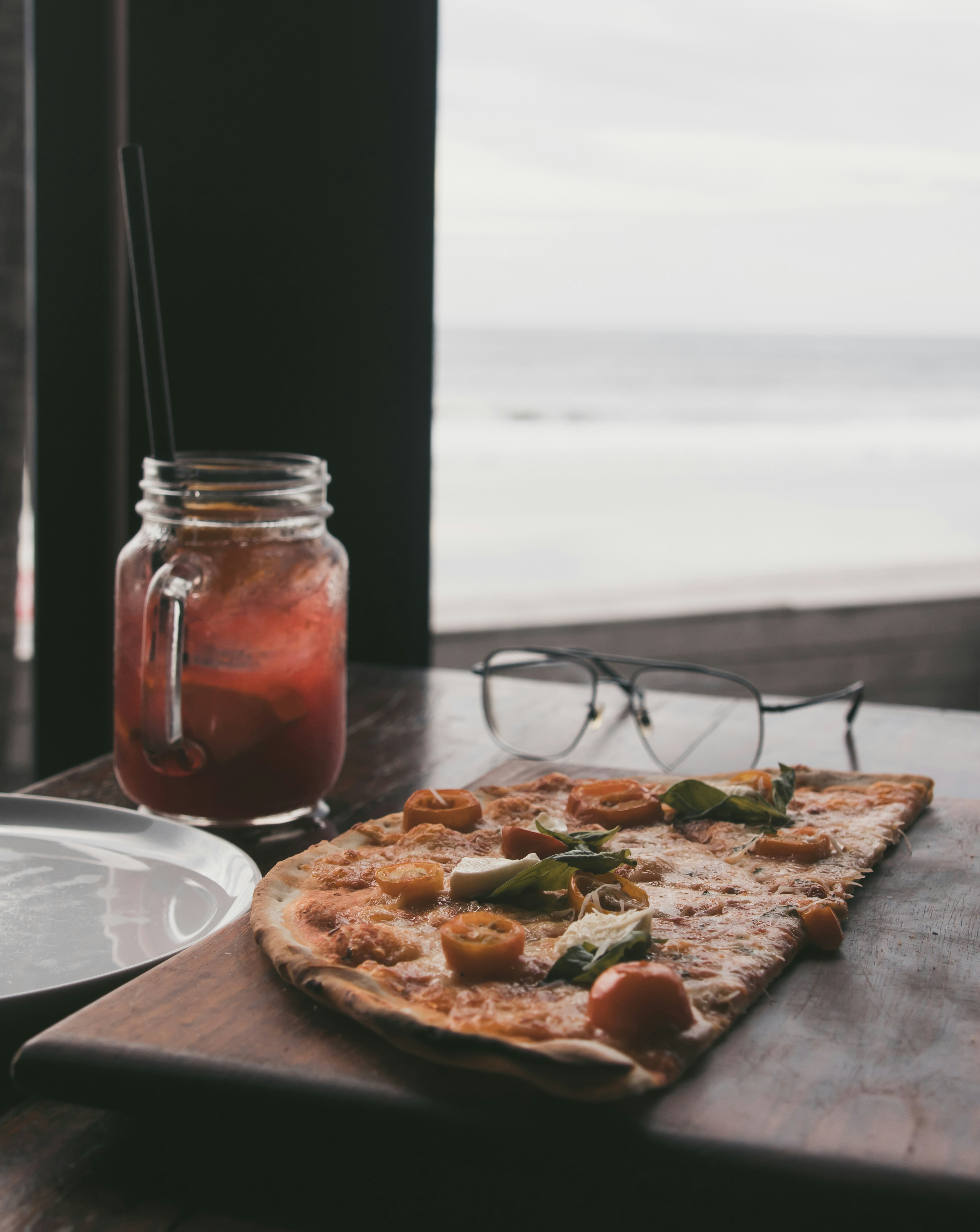 Thin crust pizza topped with fresh basil and cherry tomatoes, accompanied by a refreshing drink, set against a backdrop of the ocean.