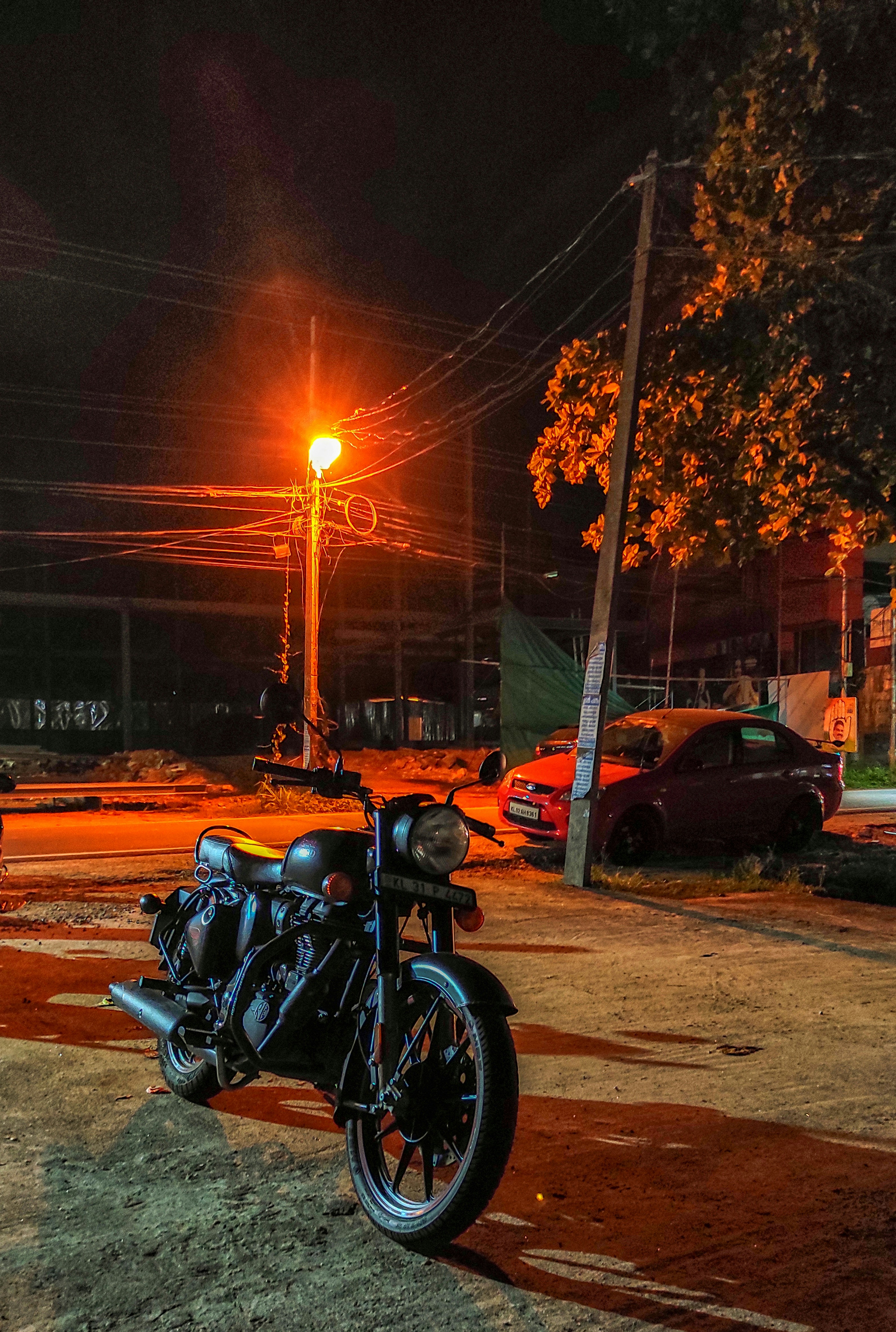 A vintage motorcycle stands against a backdrop of urban night, illuminated by a streetlight, with power lines creating a web above. The scene captures the essence of solitude in a bustling city.