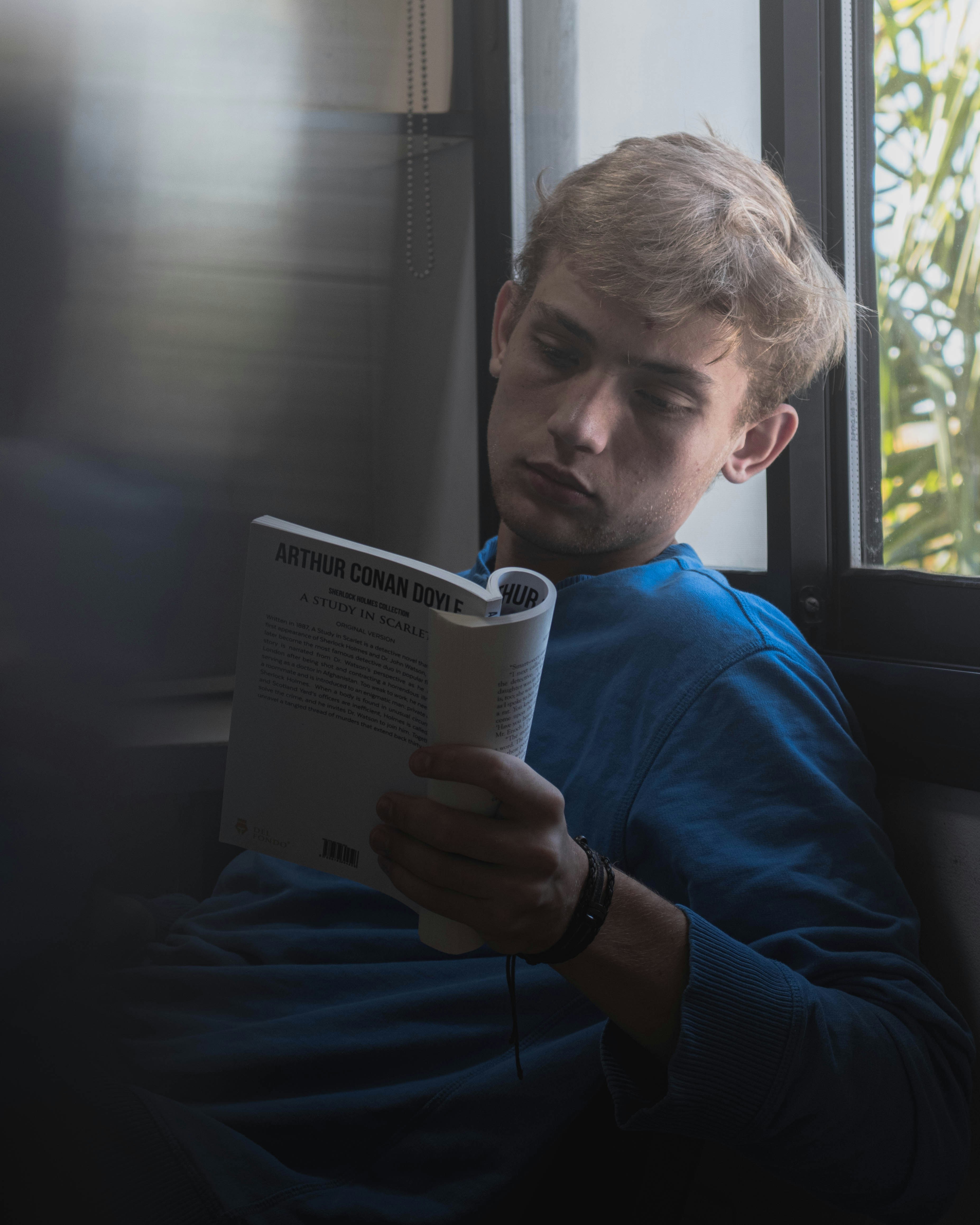 Young man absorbed in reading a book by Arthur Conan Doyle, seated by a window with soft natural light illuminating his focused expression.