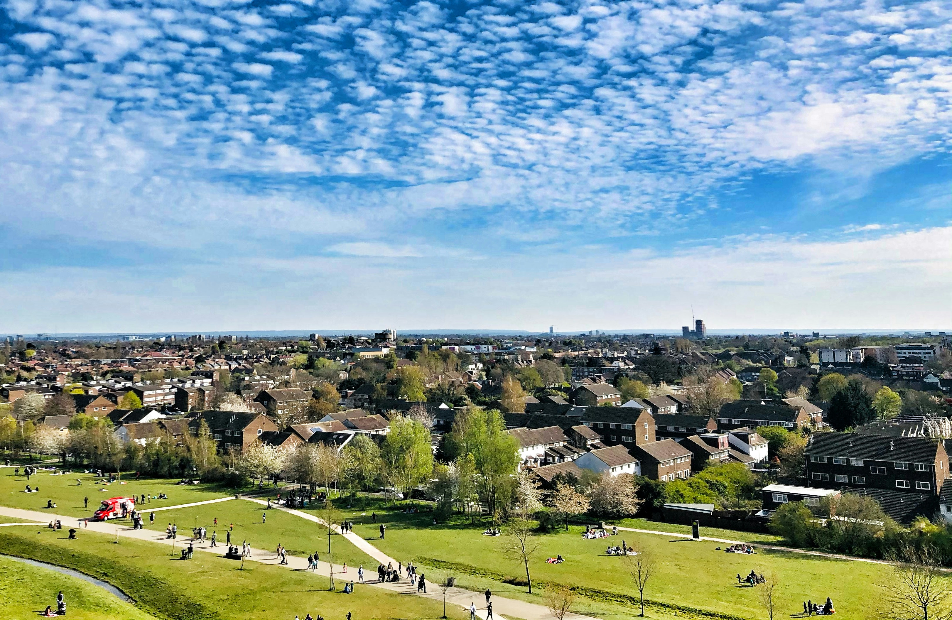 Lush green park filled with people enjoying leisure time, framed by a sprawling urban landscape and a dynamic sky. The scene showcases a harmonious blend of nature and city life.