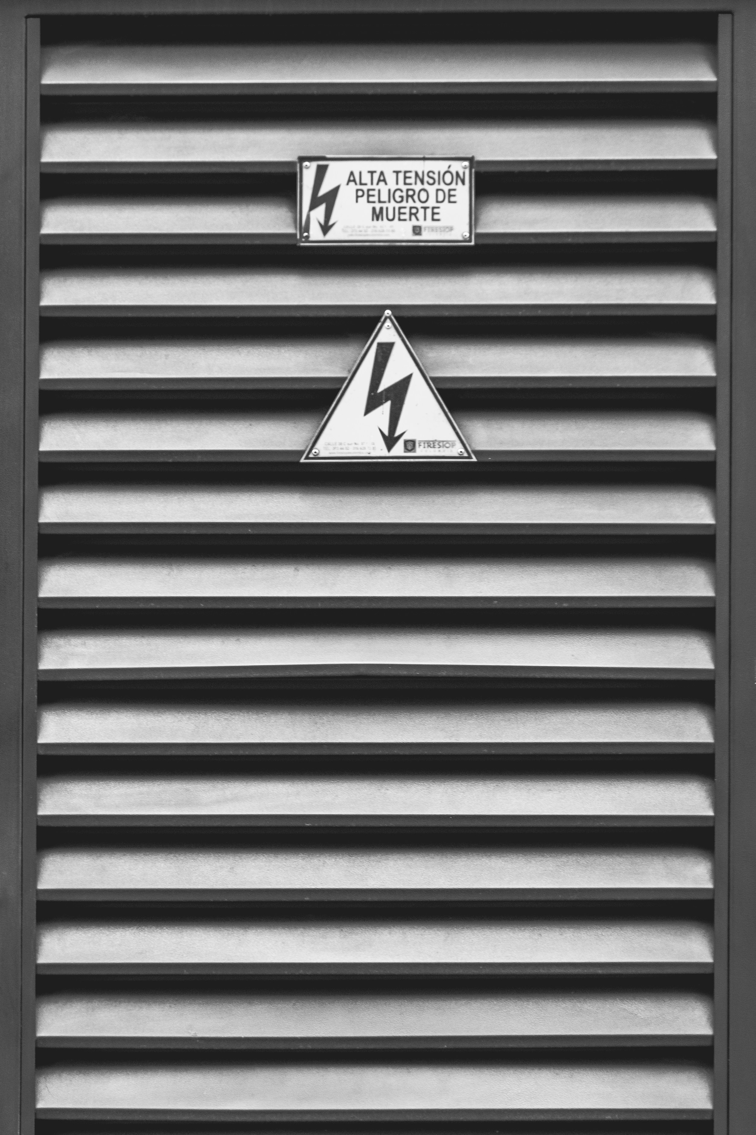 Black-and-white photograph of a metal louvered panel featuring a triangular lightning bolt warning sign and a small rectangular caution plaque.