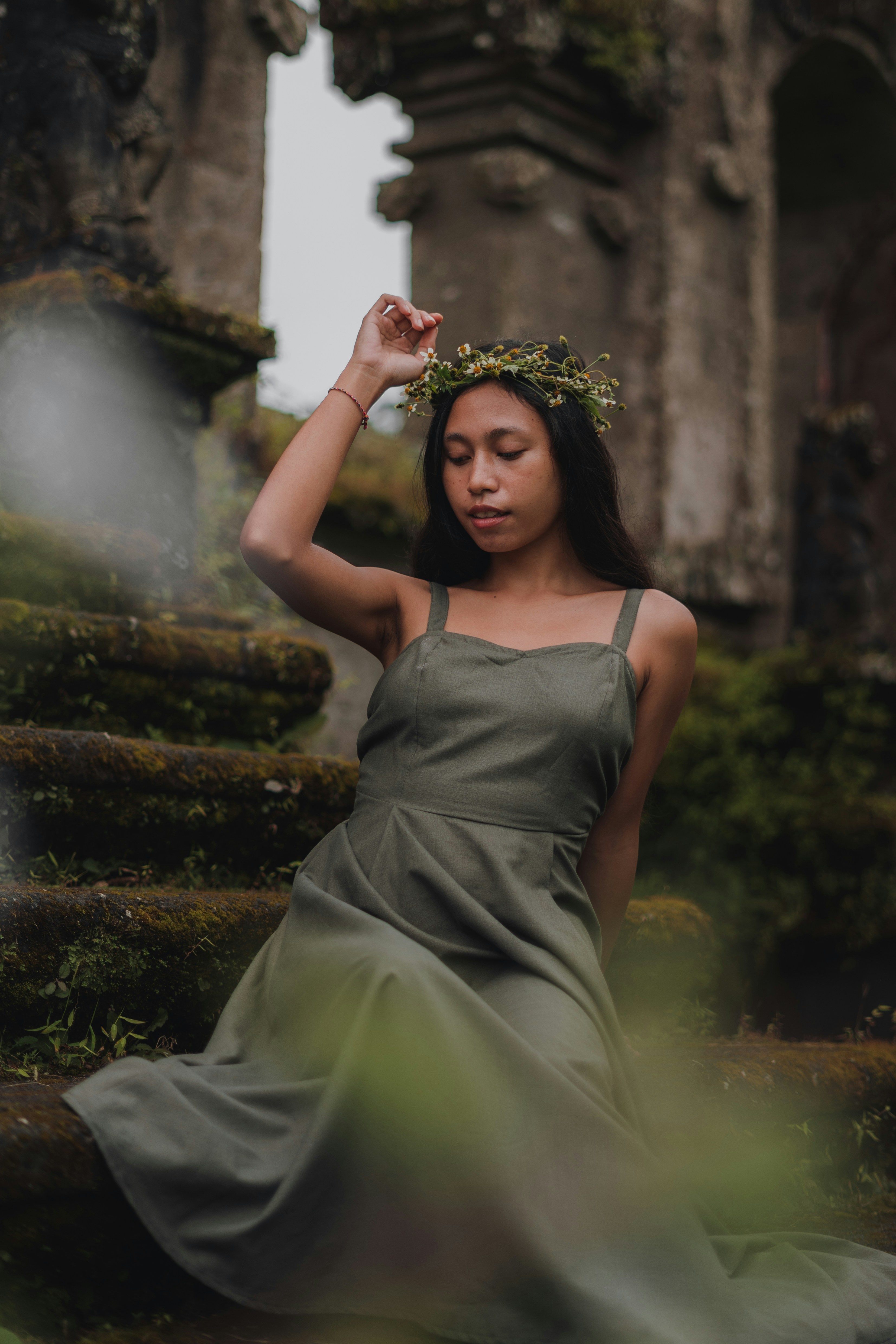 A woman adorned with a floral crown sits gracefully on moss-covered steps, exuding tranquility amidst ancient stone structures.