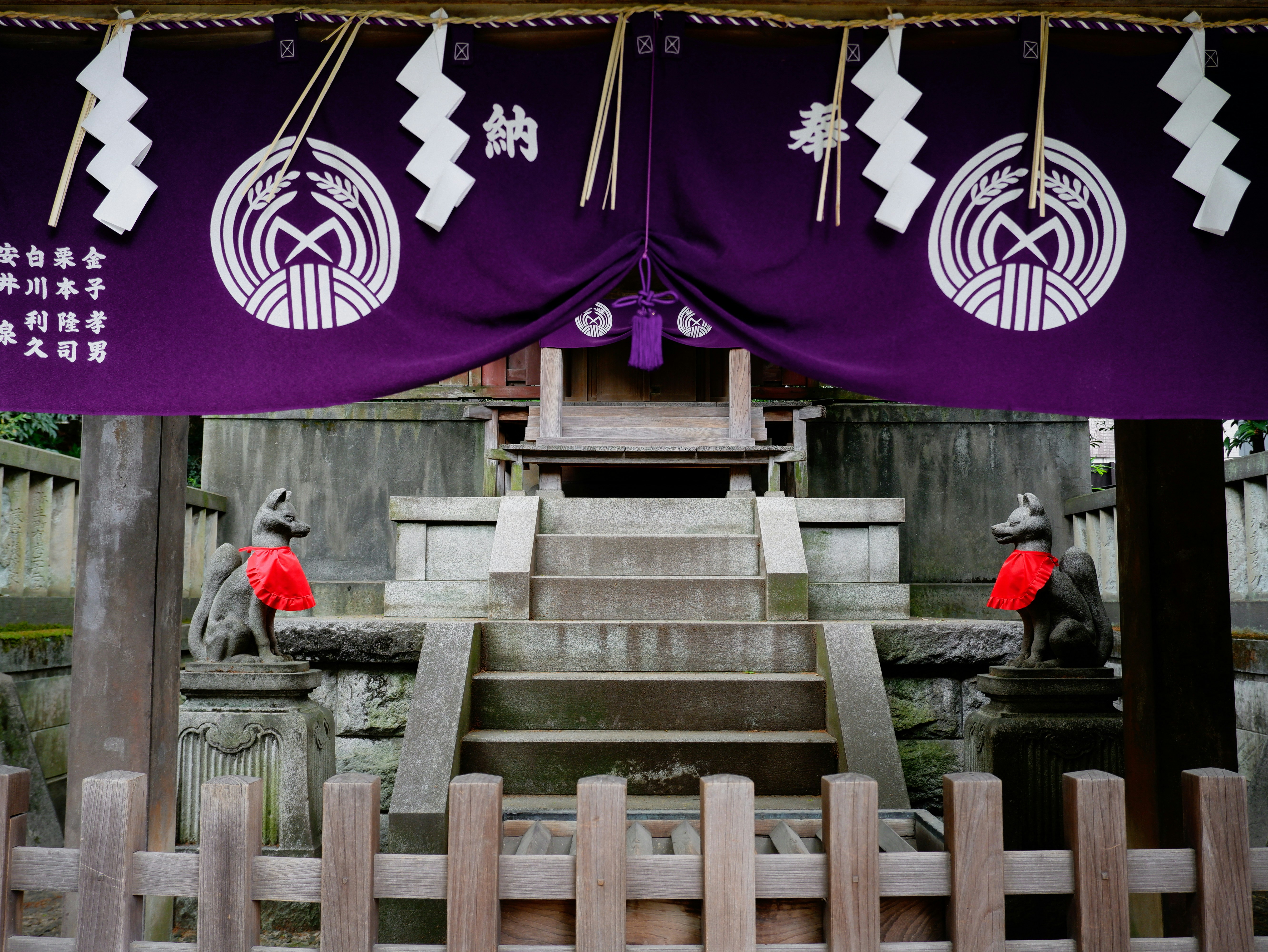 Stone steps ascend to a small shrine framed by indigo banners and guardian fox statues wearing red bibs.