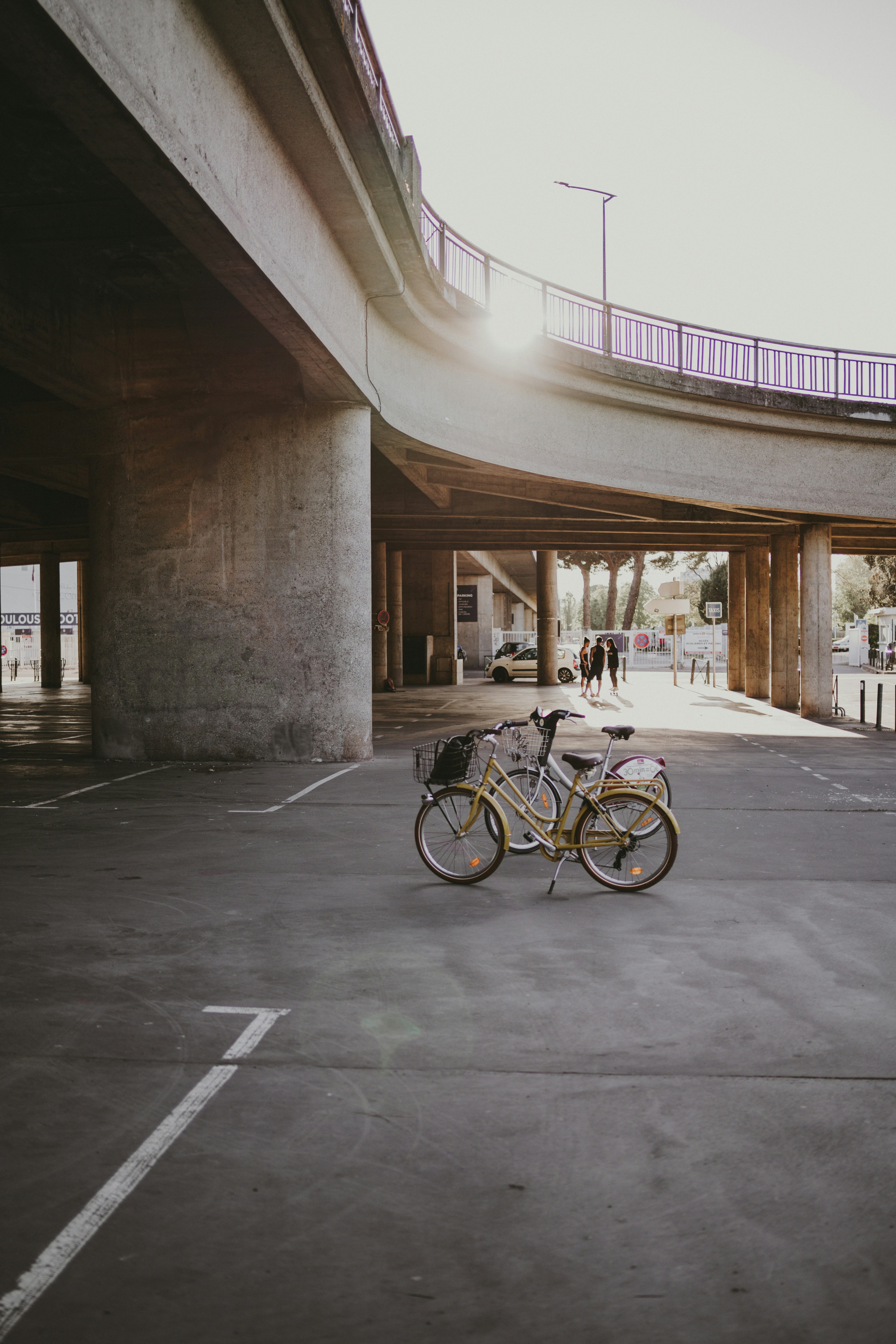 Yellow bicycle parked in a vast, empty space under a concrete overpass, with pedestrians in the distance. 